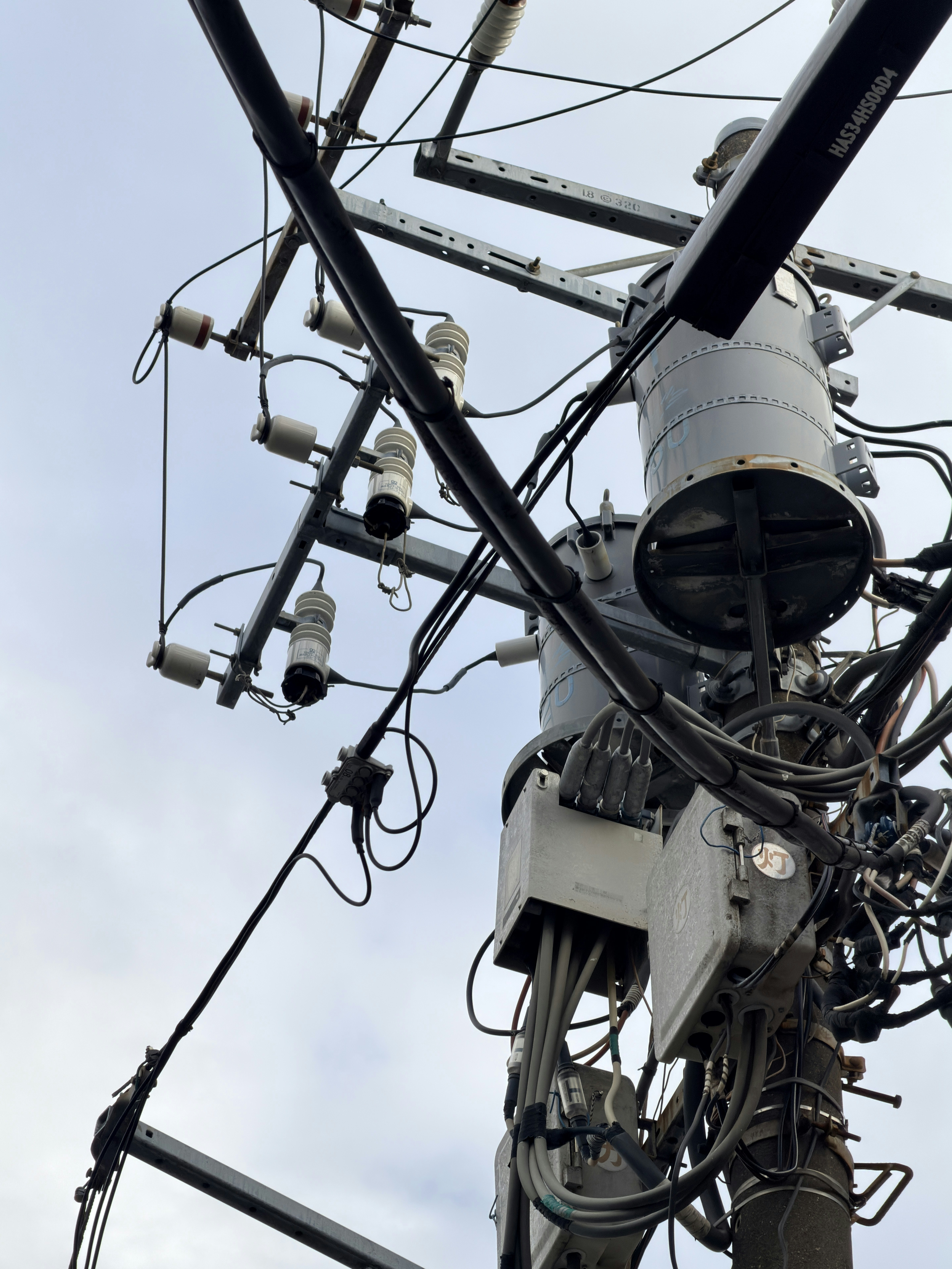 Close view of a utility pole with transformers, insulators, and a tangle of cables against a pale sky.