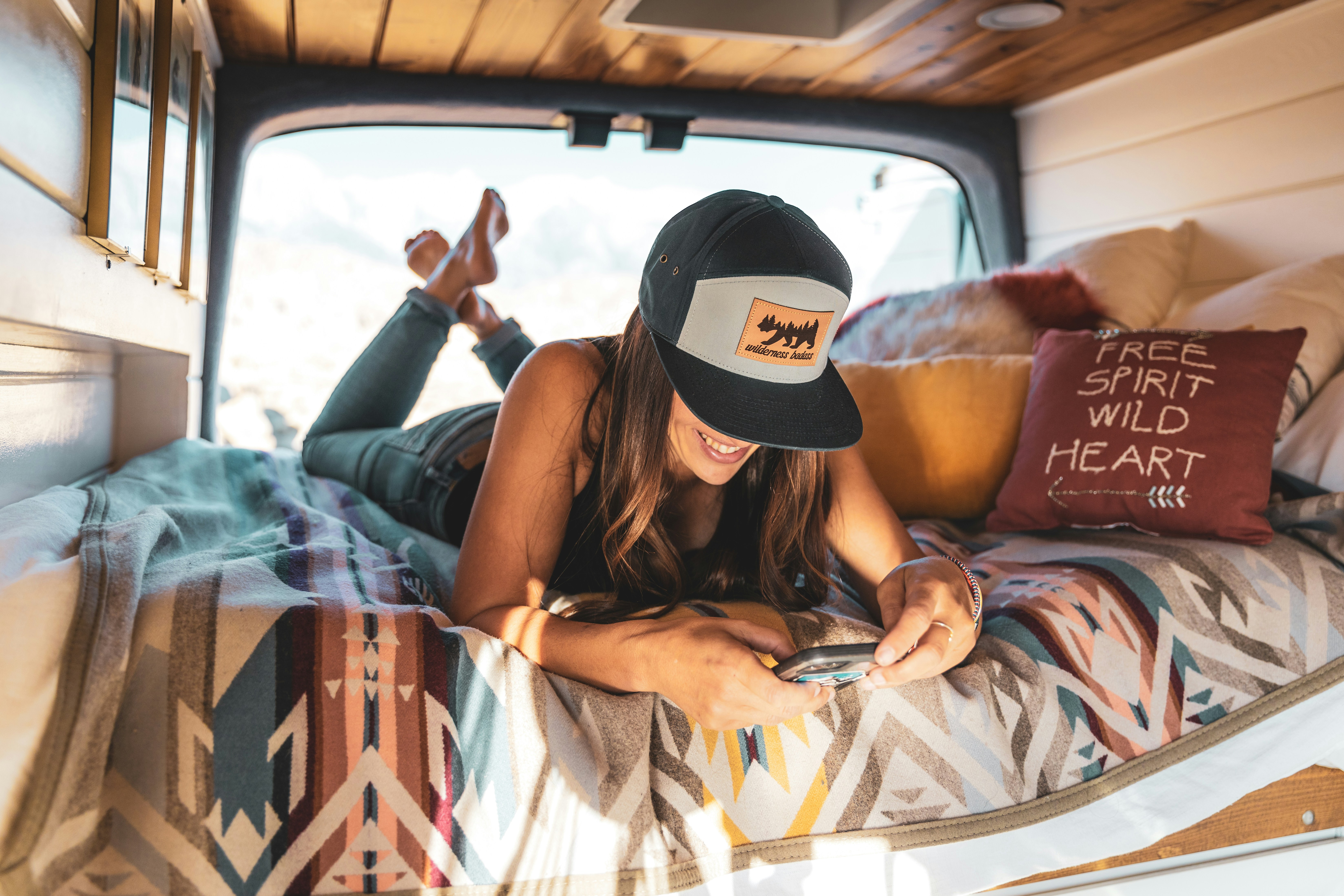 A young woman lies on a patterned blanket inside a vintage camper, focused on a handheld device, with soft natural light filling the space and a pillow reading 'FREE SPIRIT WILL HEART' in view.
