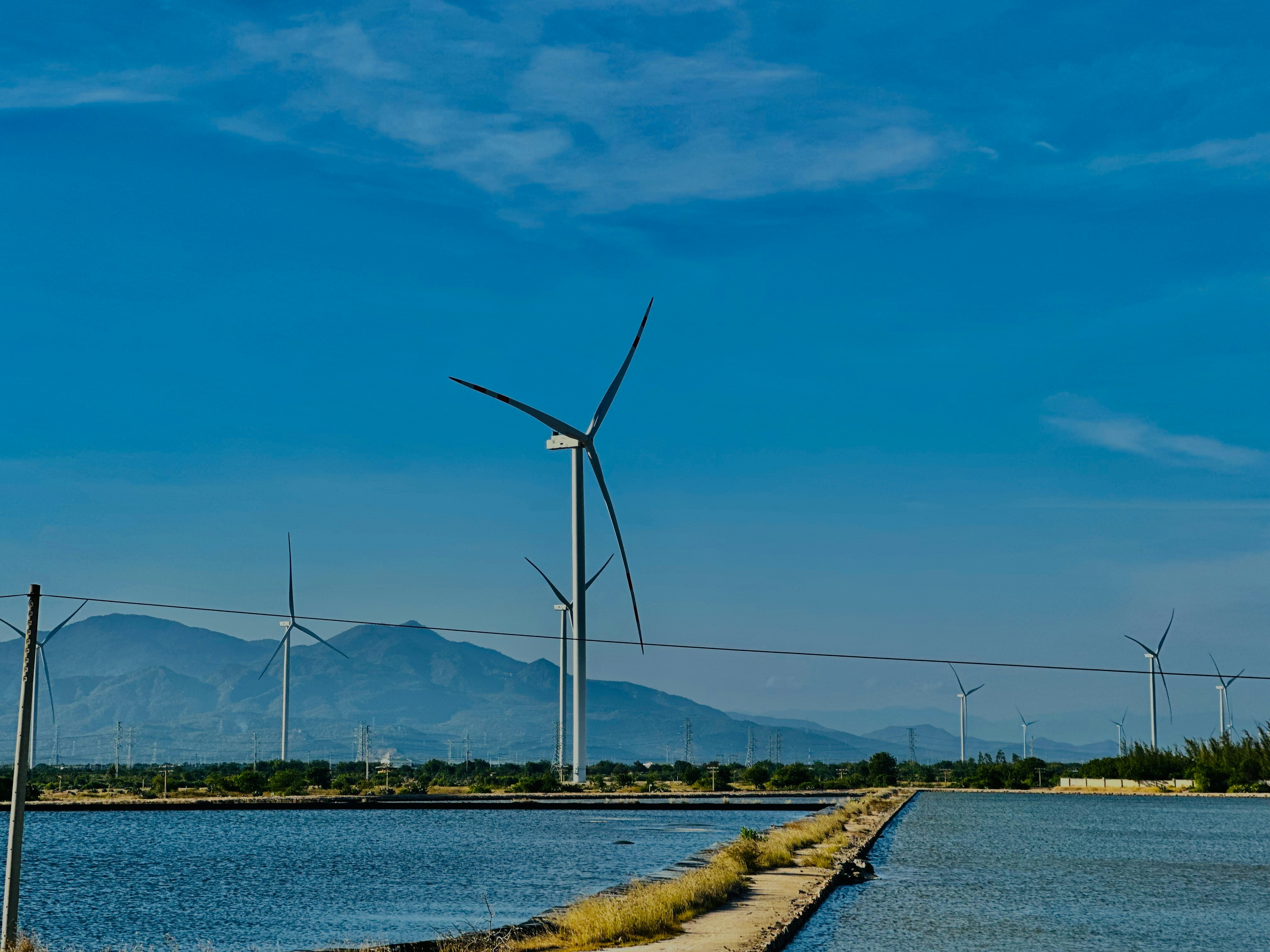 Wind turbines rise above a low-lying landscape with a calm water canal in the foreground and distant mountains under a clear blue sky.