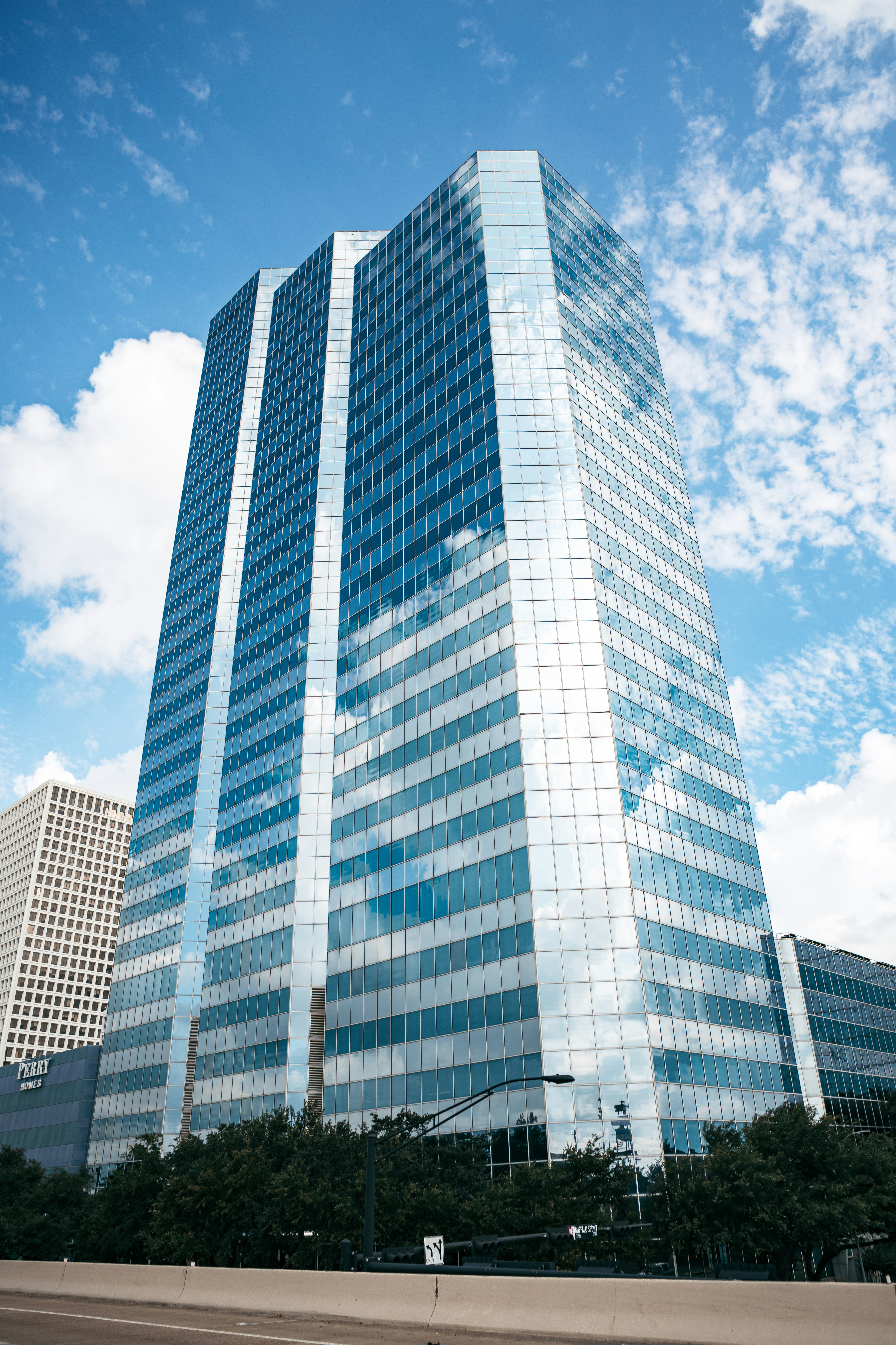 Sleek glass skyscraper rising against a bright blue sky with scattered clouds, reflected windows creating geometric patterns. A modern cityscape view emphasizing architectural symmetry and vertical scale.