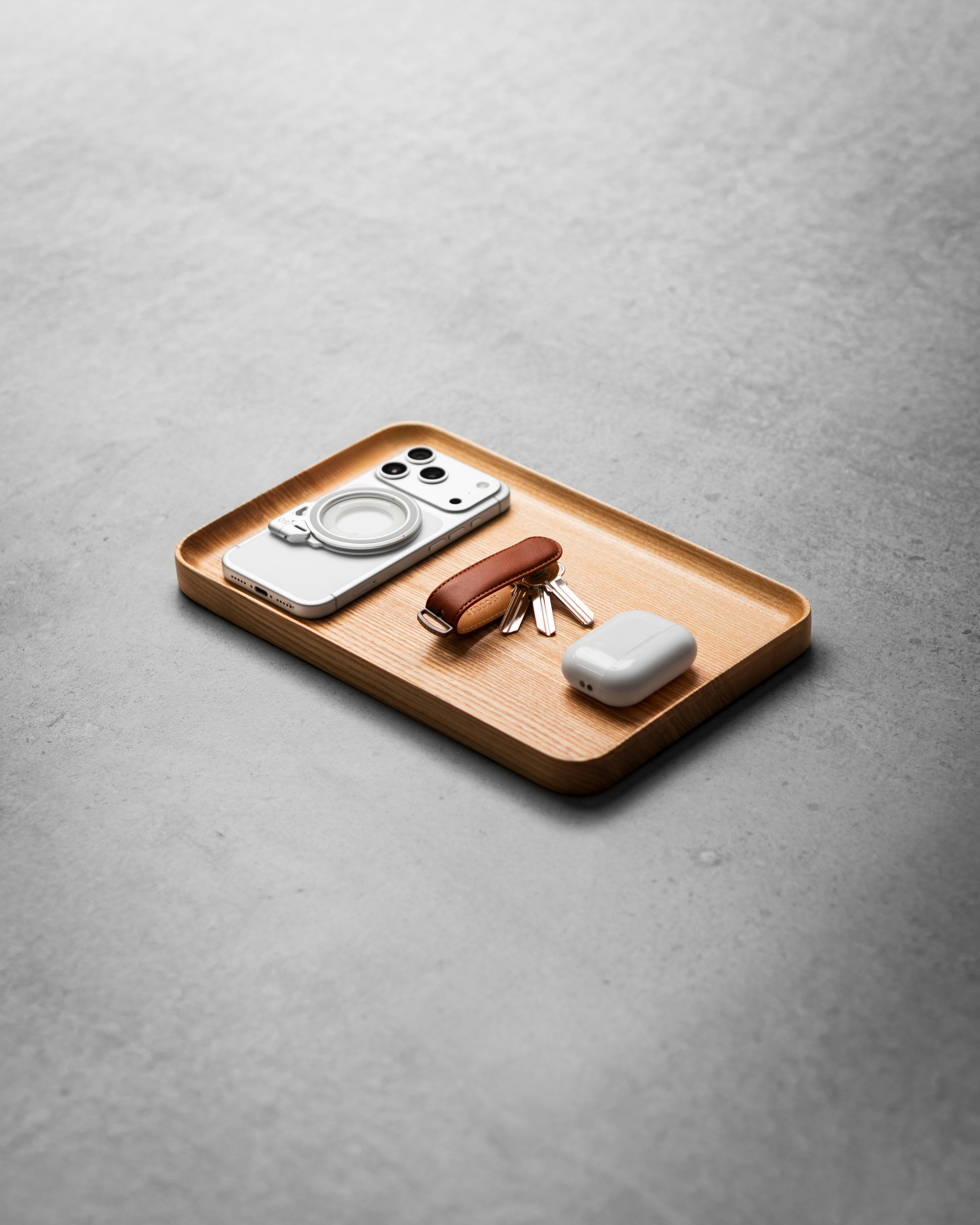 Minimal desk scene with a wooden tray holding a sleek white device, a brown key cover, and a small white case on a smooth gray surface.