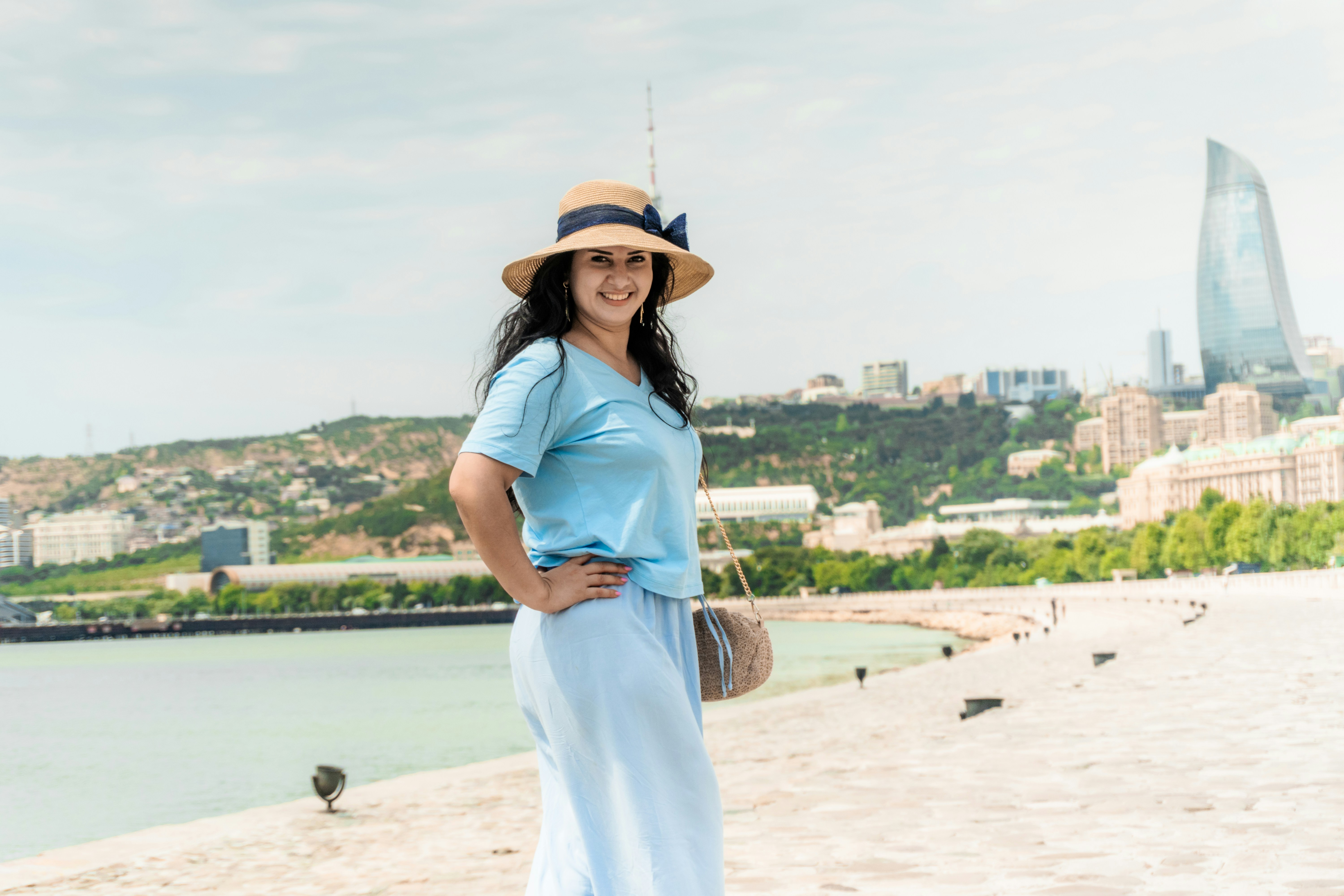 Coastal Confidence: A woman in a light blue dress on a sunlit beach with a cityscape backdrop