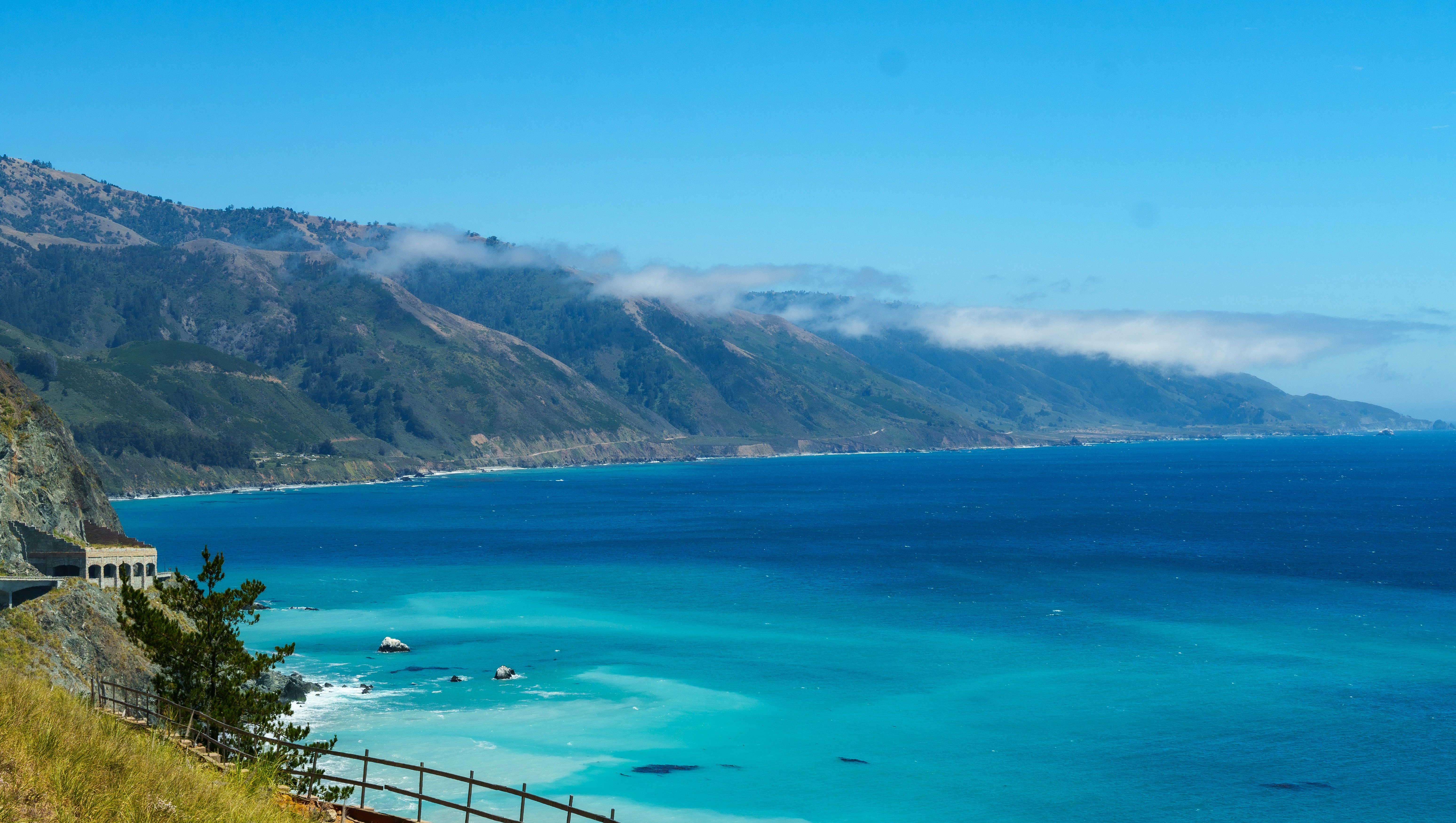 Vibrant coastal scene along a cliffside road with turquoise water and distant mountains under a clear blue sky. A subtle railing lines the foreground, guiding the eye toward the expansive sea and horizon.