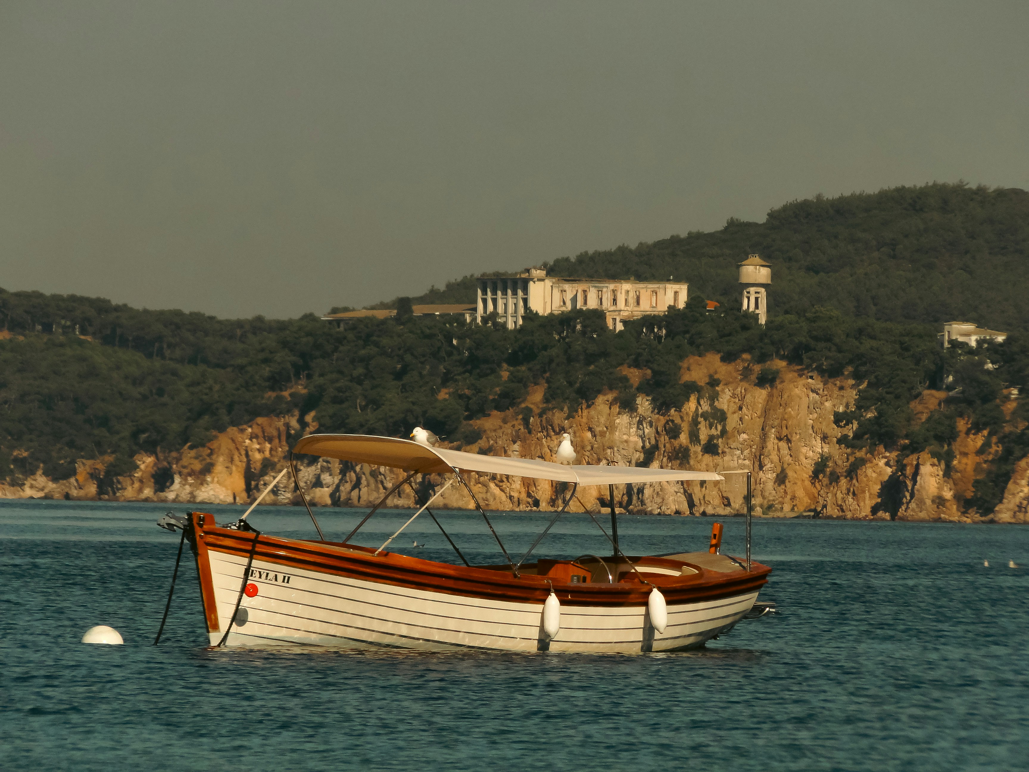 A small wooden boat rests on calm sea near a rocky coastline under muted afternoon light, with a distant hill and buildings in the background.
