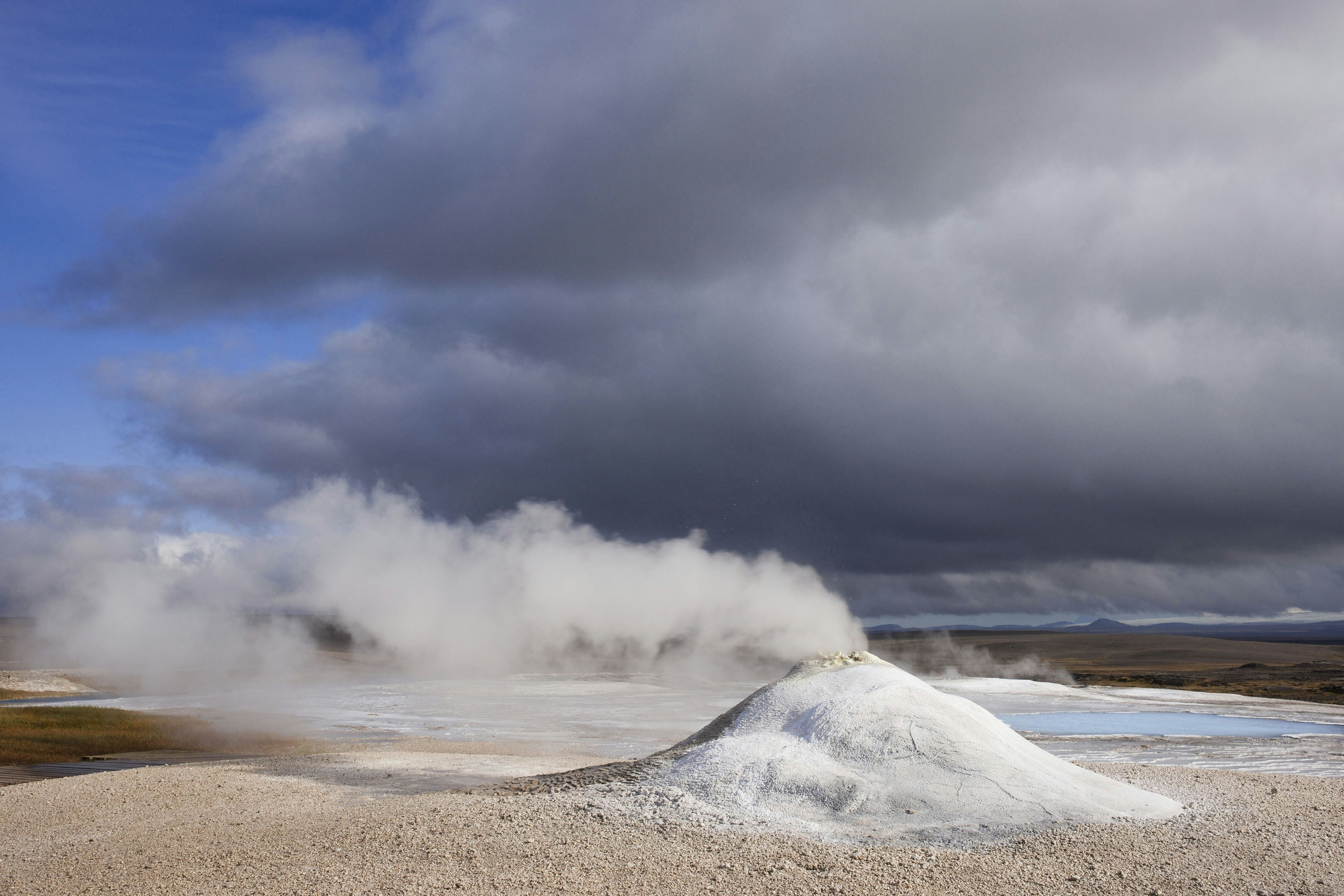 Steam plume from a mineral cone rises over pale terrain with a distant horizon under dramatic clouds.