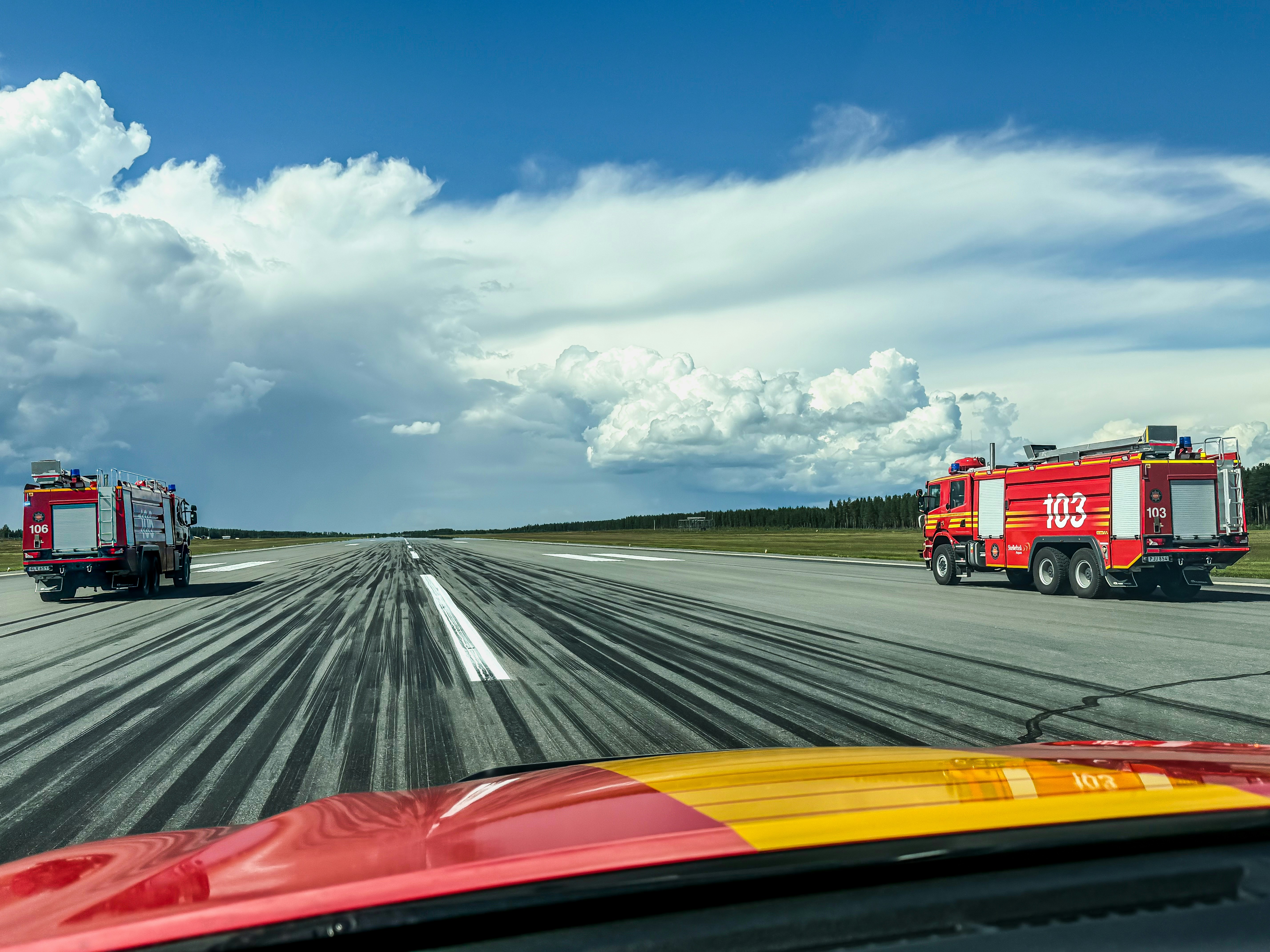 Toward the horizon, two fire trucks sit ready on a runway as the driver's viewpoint shows the striped centerline and colorful hood in the foreground.