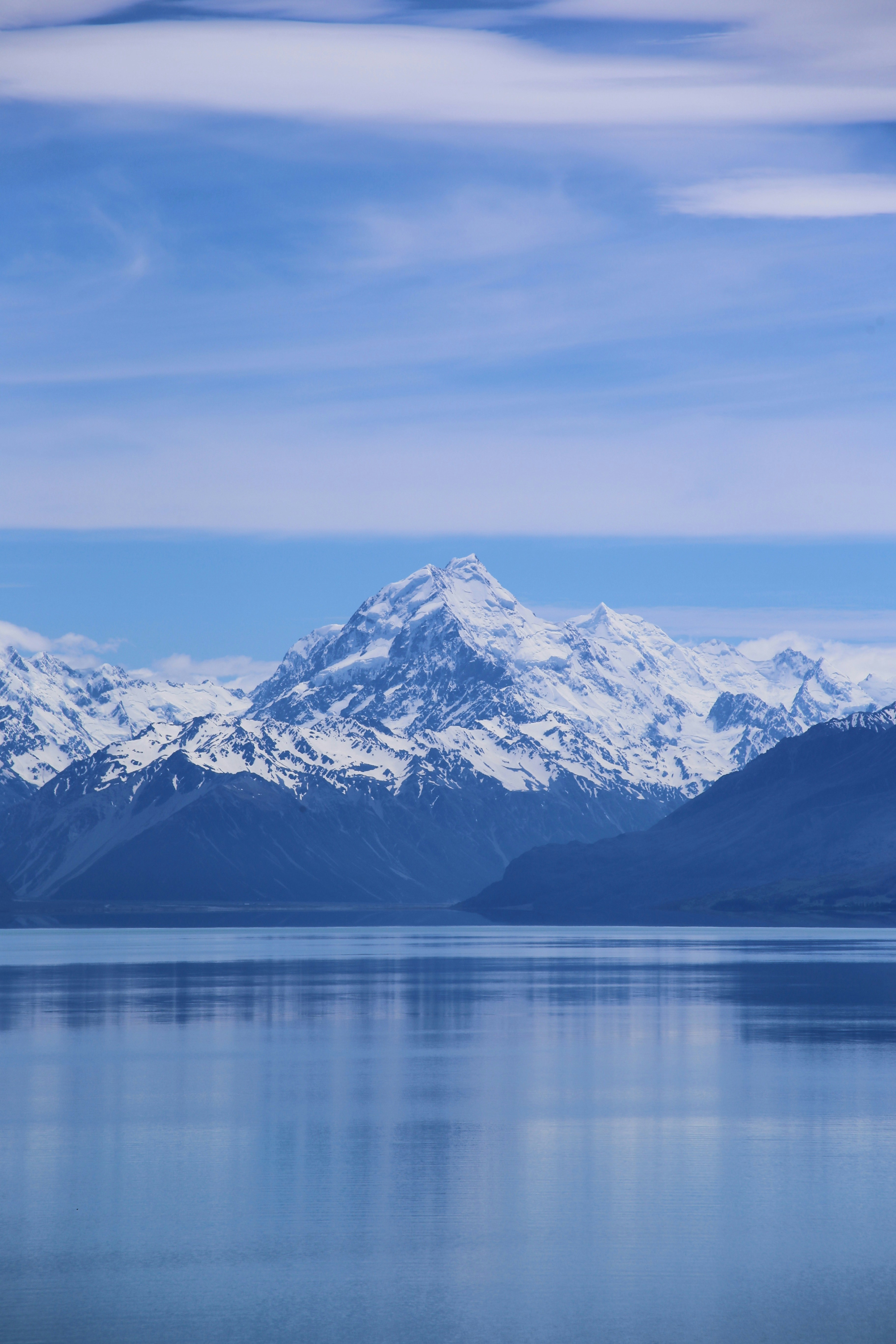 Snow-capped mountains rise over a calm, glassy fjord under a soft blue sky, with cool tones and pristine reflections. A serene landscape emphasizing vast distance and natural symmetry.