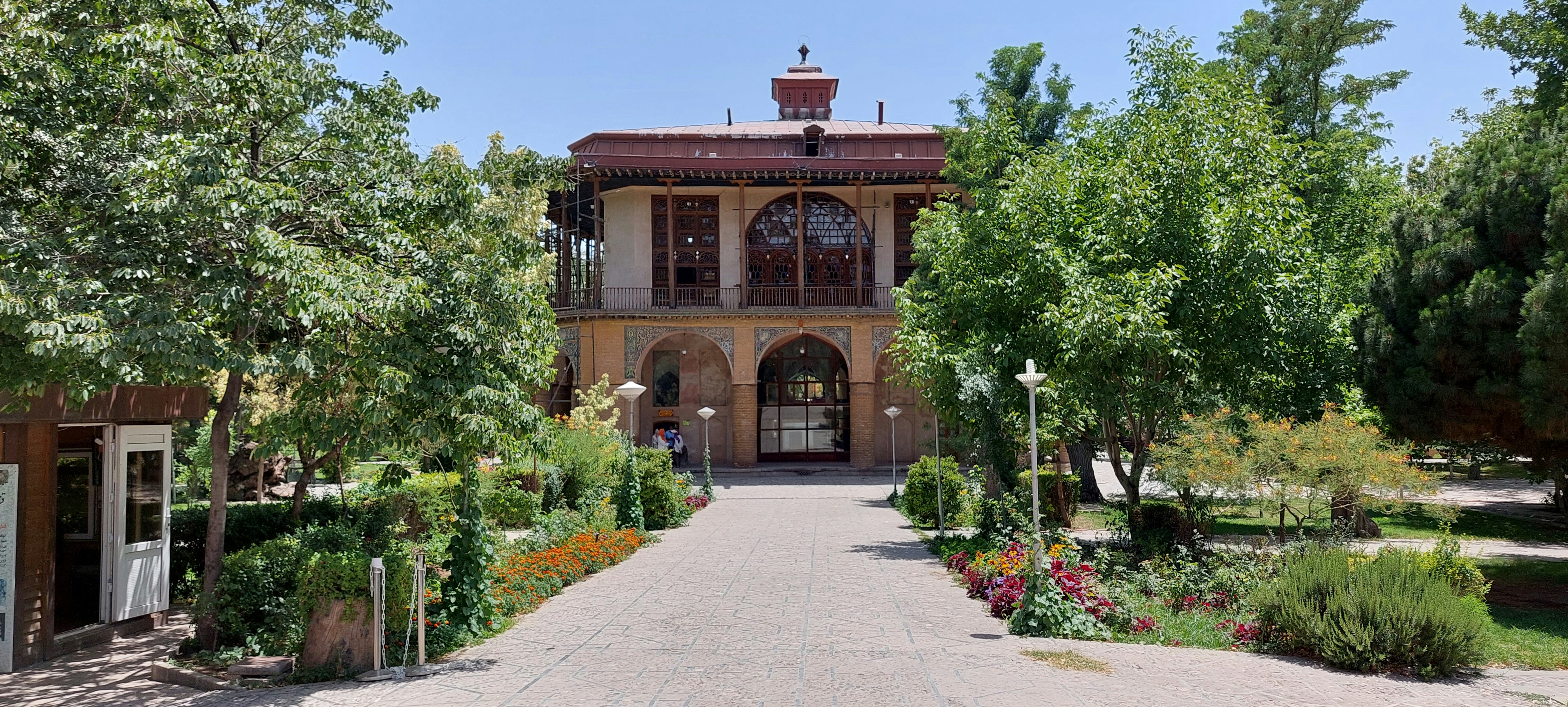Sunlit historic building framed by a manicured garden and paved path, inviting entry through an arched facade; a serene courtyard scene with greenery and colorful blooms.