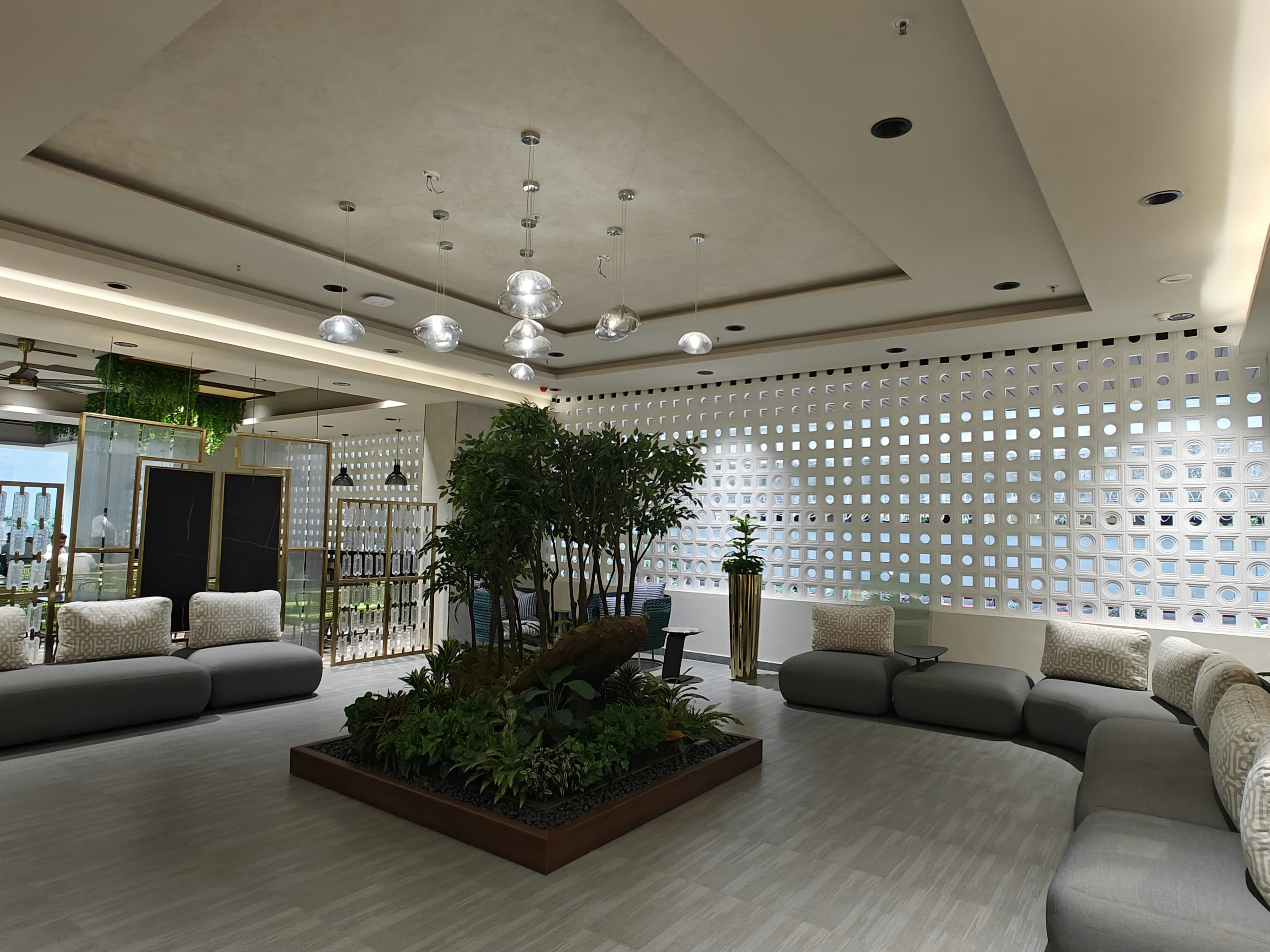 Contemporary hotel lounge with a central planter and surrounding modular seating, glass blocks along a sunlit wall, and decorative ceiling fixtures.