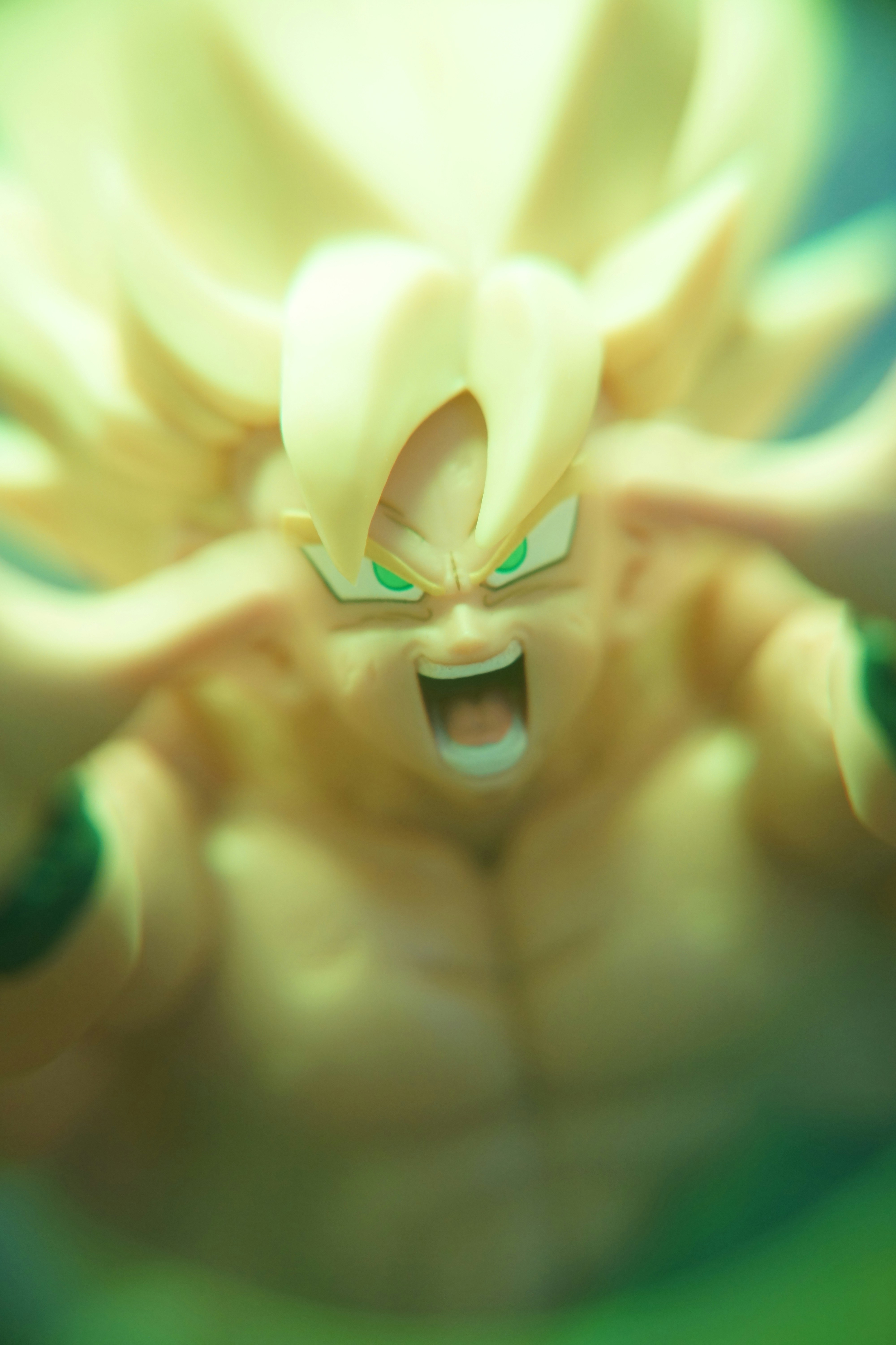 A close-up shot of a Dragon Ball character figure in Super Saiyan form, dramatic mouth open as if shouting, with soft green-yellow bokeh backdrop.