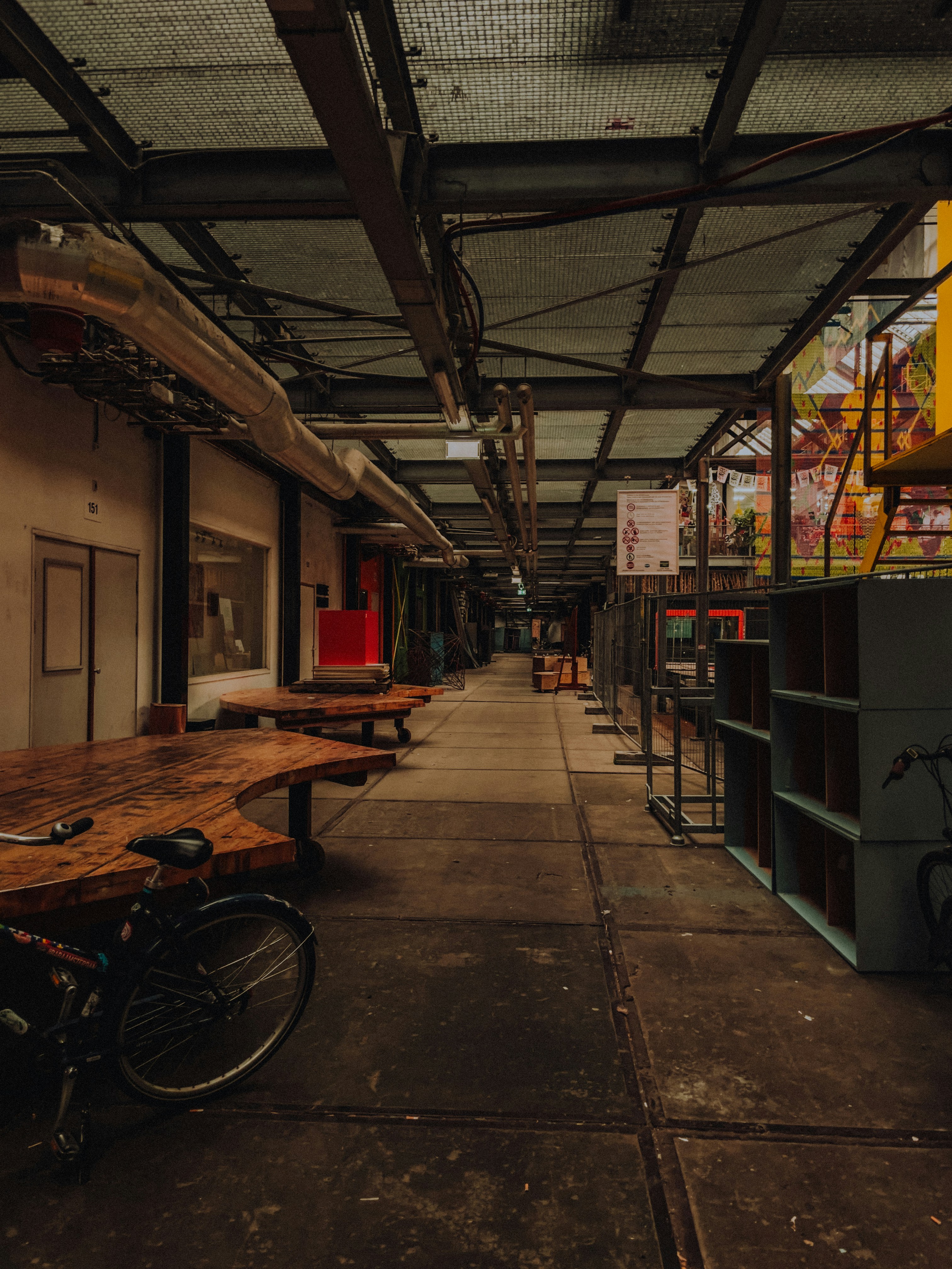 Dimly lit market corridor with long perspective, bicycles and wooden tables along the left, shelving on the right, and a colorful mural visible in the far distance.