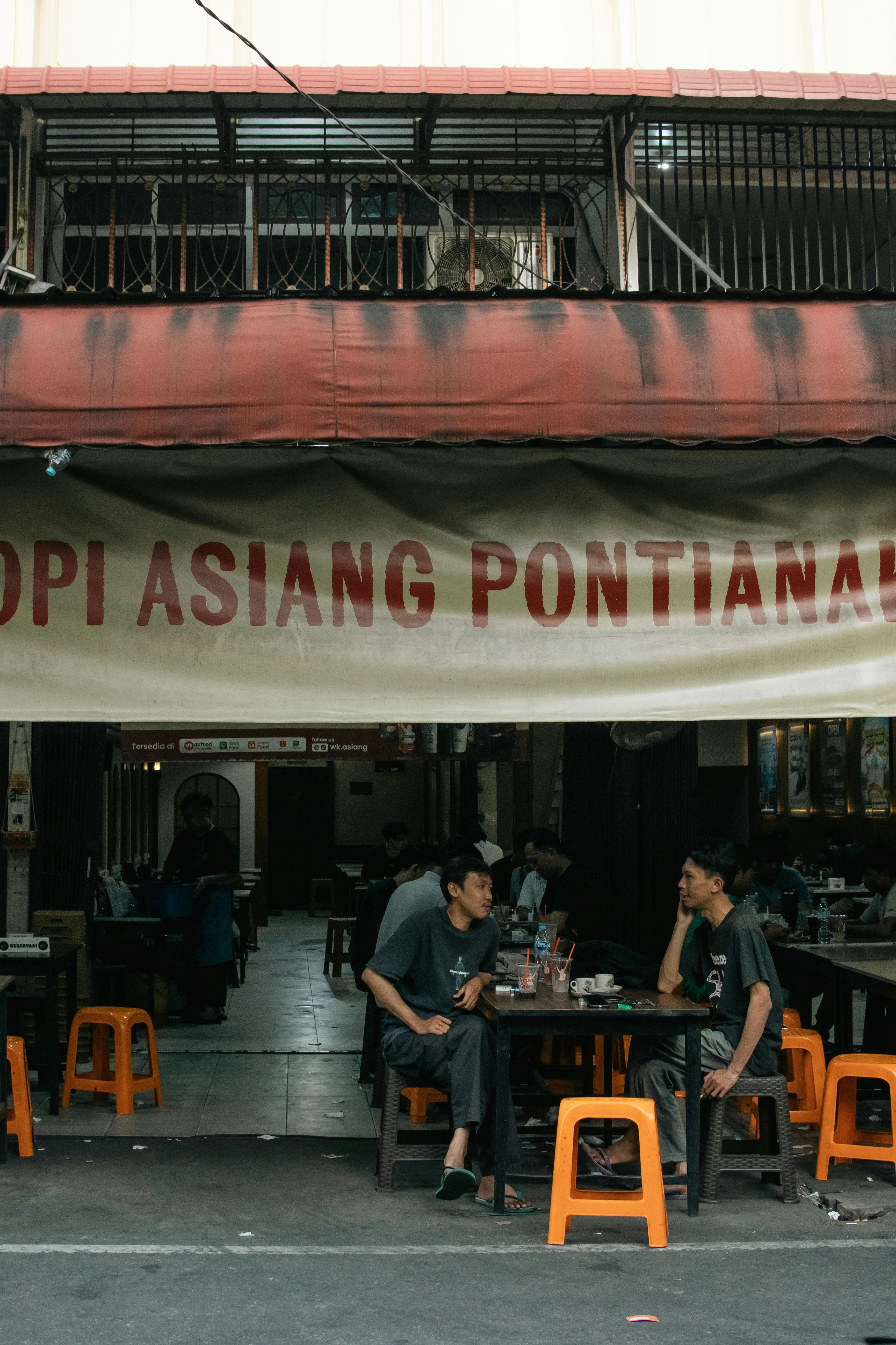 Two men sit and chat at a street-side cafe beneath a large banner in an urban market scene. An orange stool adds a pop of color in the foreground.