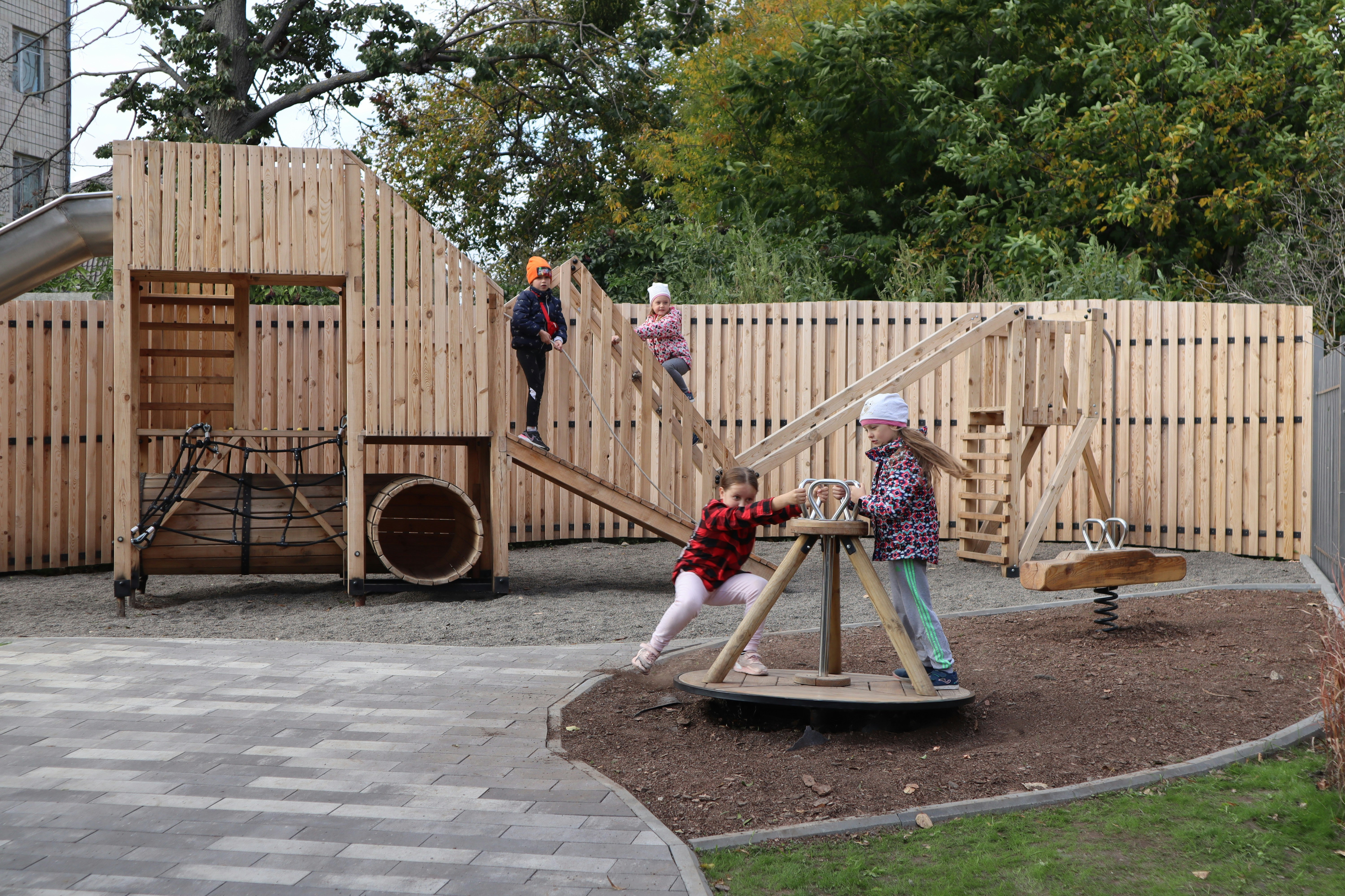 Children on a seesaw and climbing structures at a wooden playground with a paved path in the foreground.