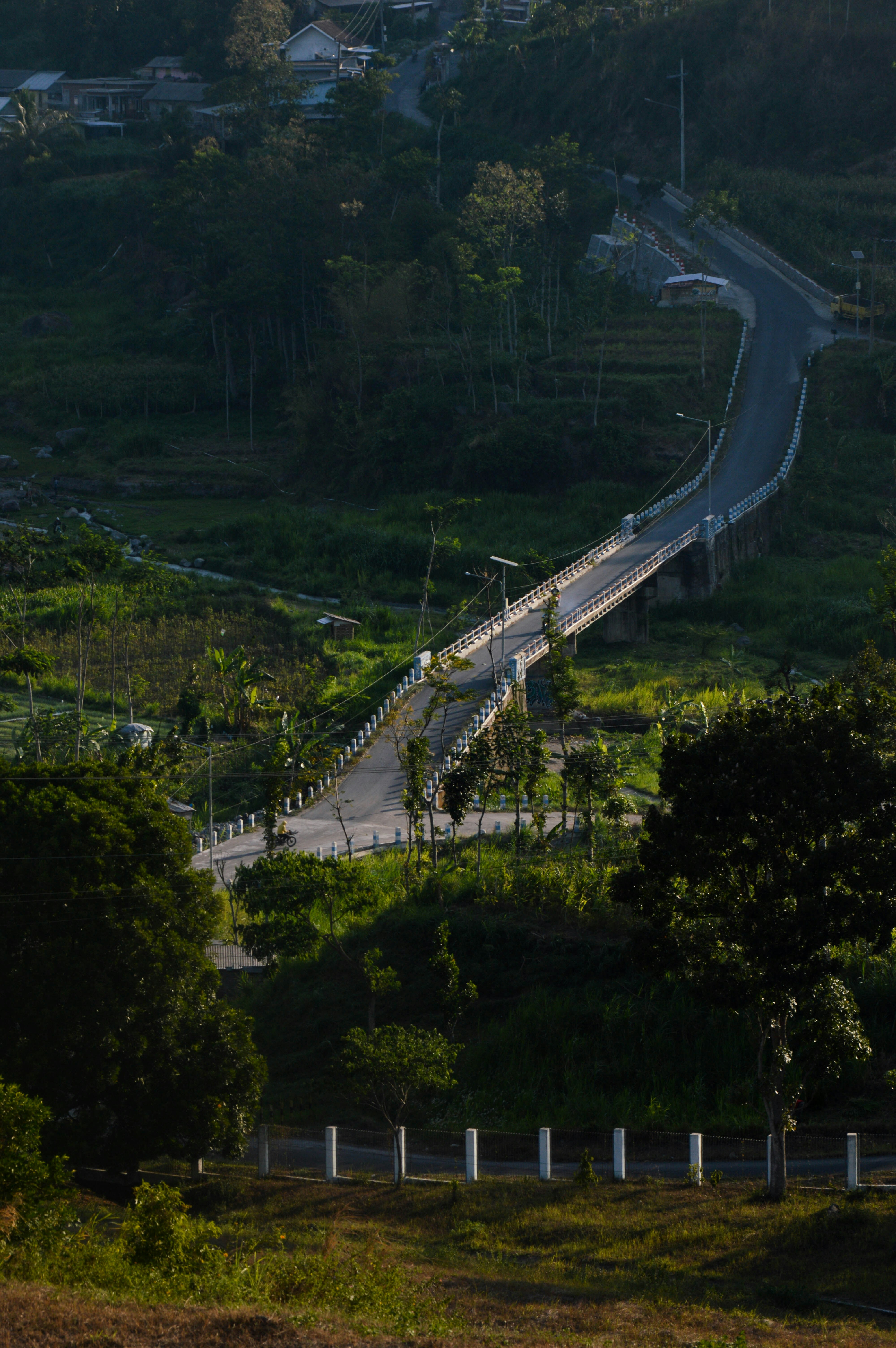 A scenic aerial view of a winding rural road crossing a bridge through lush green countryside during golden hour. Surrounded by trees, farmland, and rolling hills, the peaceful landscape highlights rural infrastructure, transportation, and the natural beauty of a tropical region.