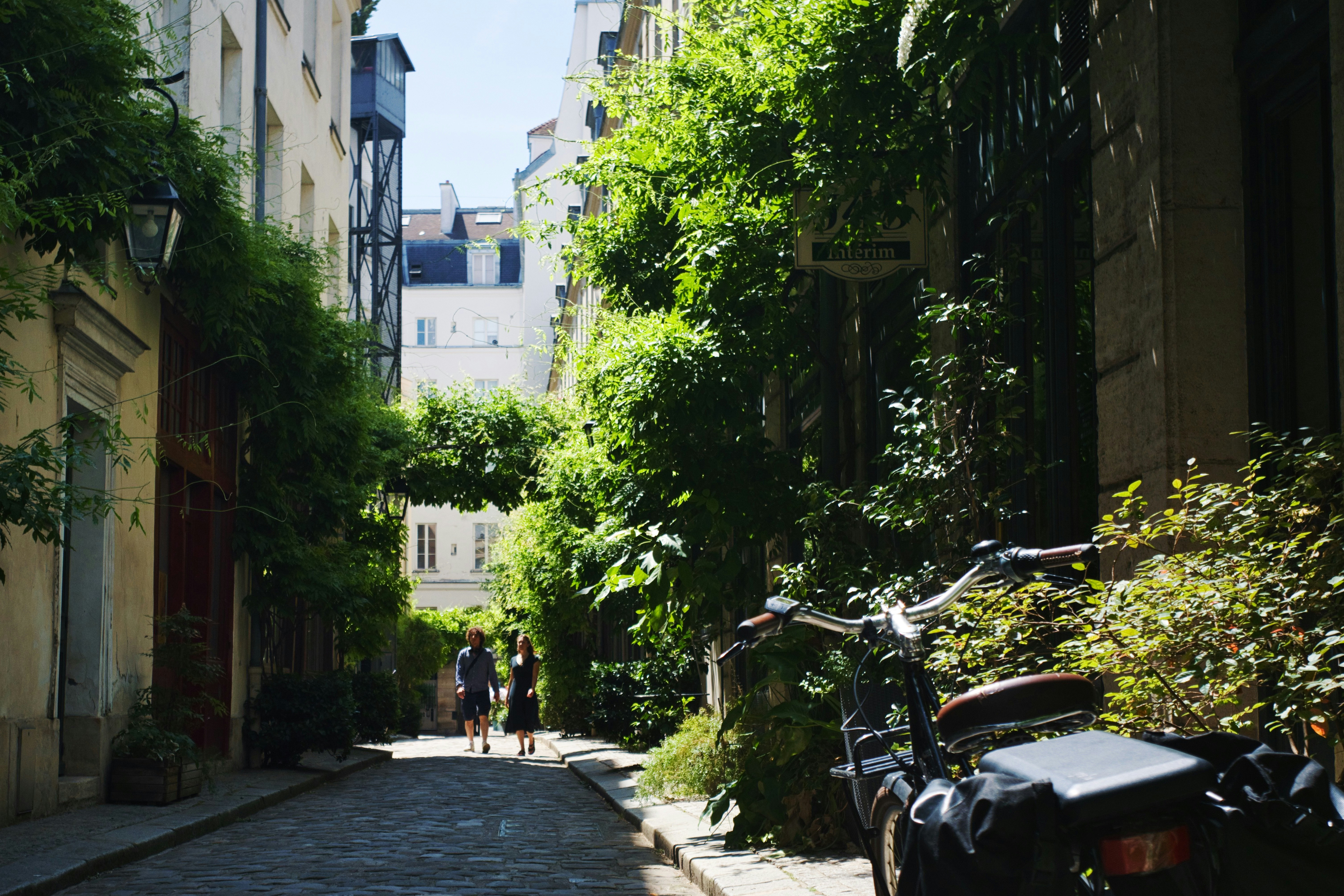 Narrow sunlit street with stone pavement, two pedestrians near the end, lush greenery on both sides, a bicycle in the foreground on the right. A tranquil urban courtyard vibe with soft daylight.