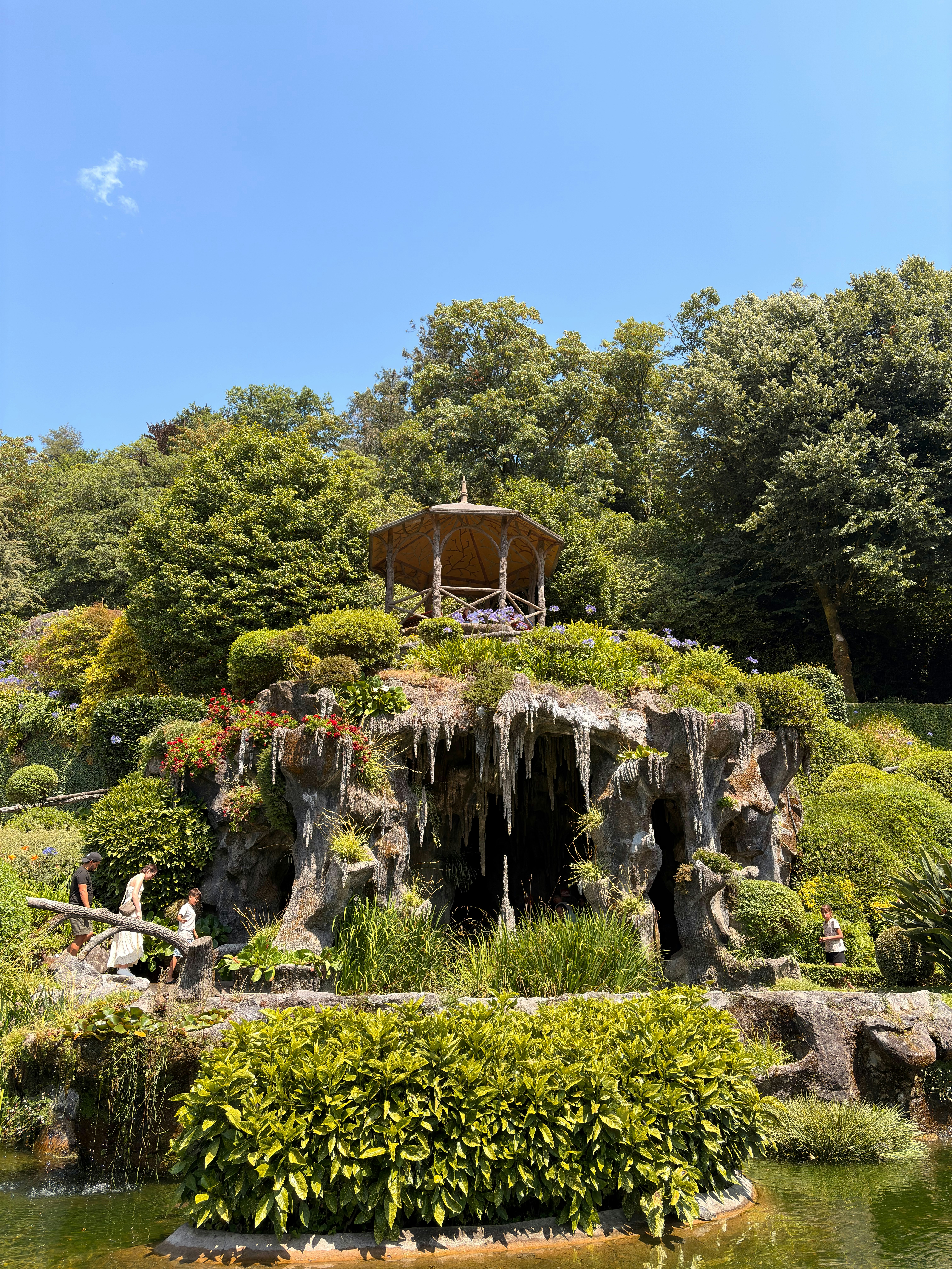 A landscaped garden with a wooden pavilion perched atop a rocky, cascading formation surrounded by lush shrubs and a small pond.