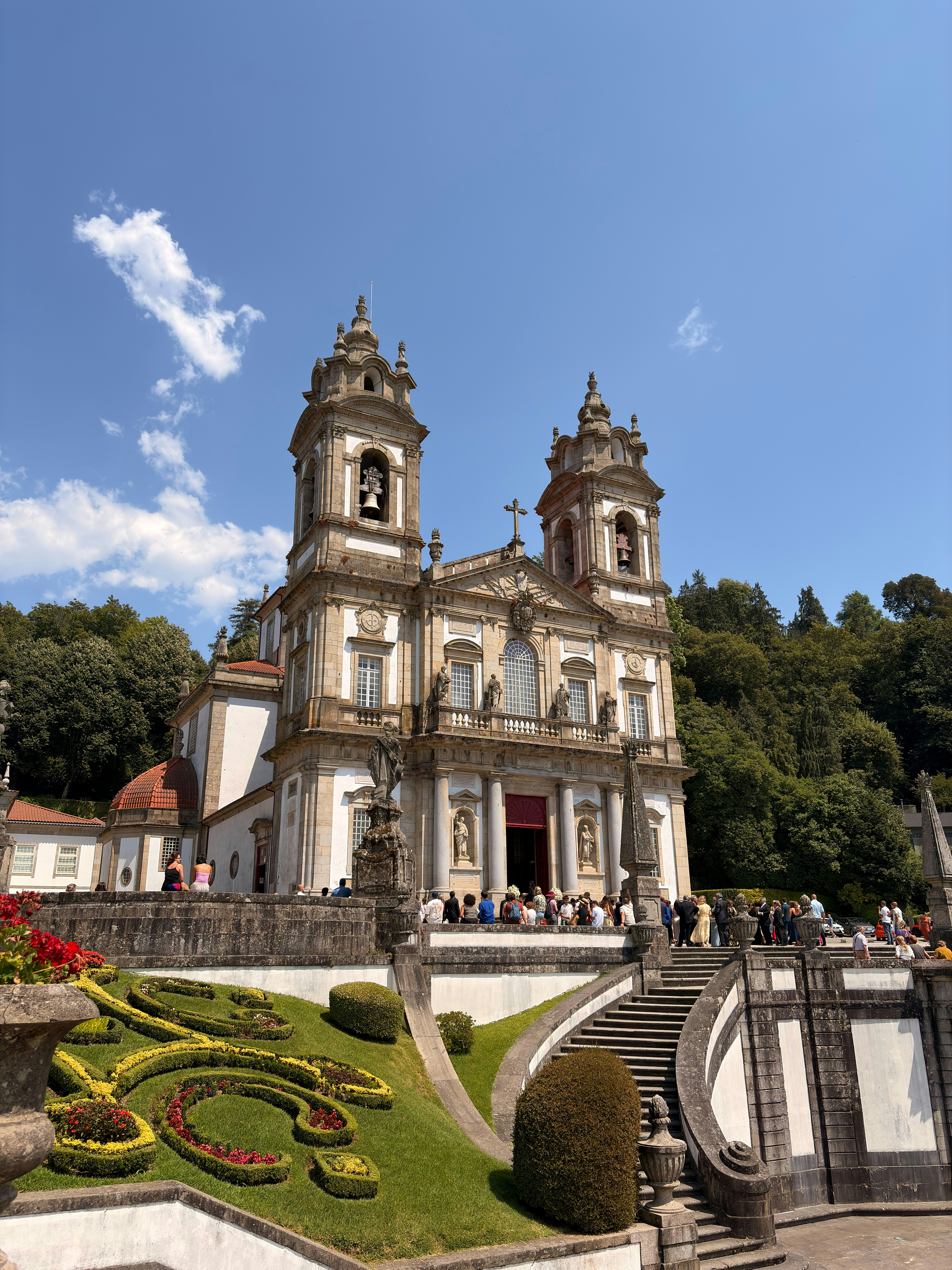 Stone church facade with twin towers and ornate details, crowds at the entrance, manicured garden terraces in the foreground under a clear blue sky. The scene captures a historical site with visitors exploring the exterior.