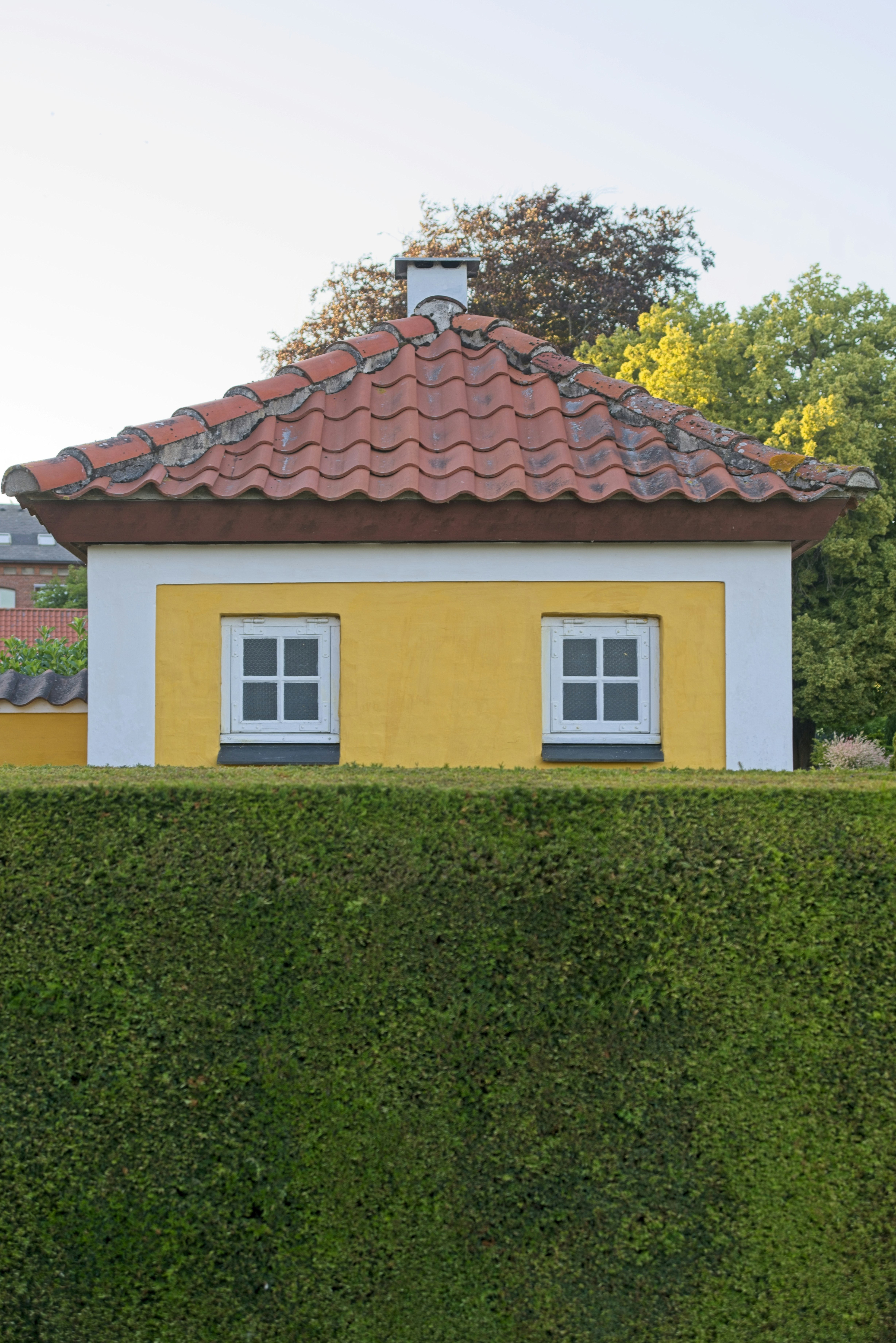 A small building adjecant to Præstø church, with symmetrical white-framed windows peeks just above a neatly trimmed hedge, its terracotta roof and squat white chimney standing out against a soft, hazy sky. Trees in the background show subtle hints of early autumn color mixed with summer green, adding depth to the simple composition. The tidy hedge line and minimalist framing give the scene a clean, almost graphic quality. This charming little building captures a quiet, understated corner of Præstø on a hot July evening.