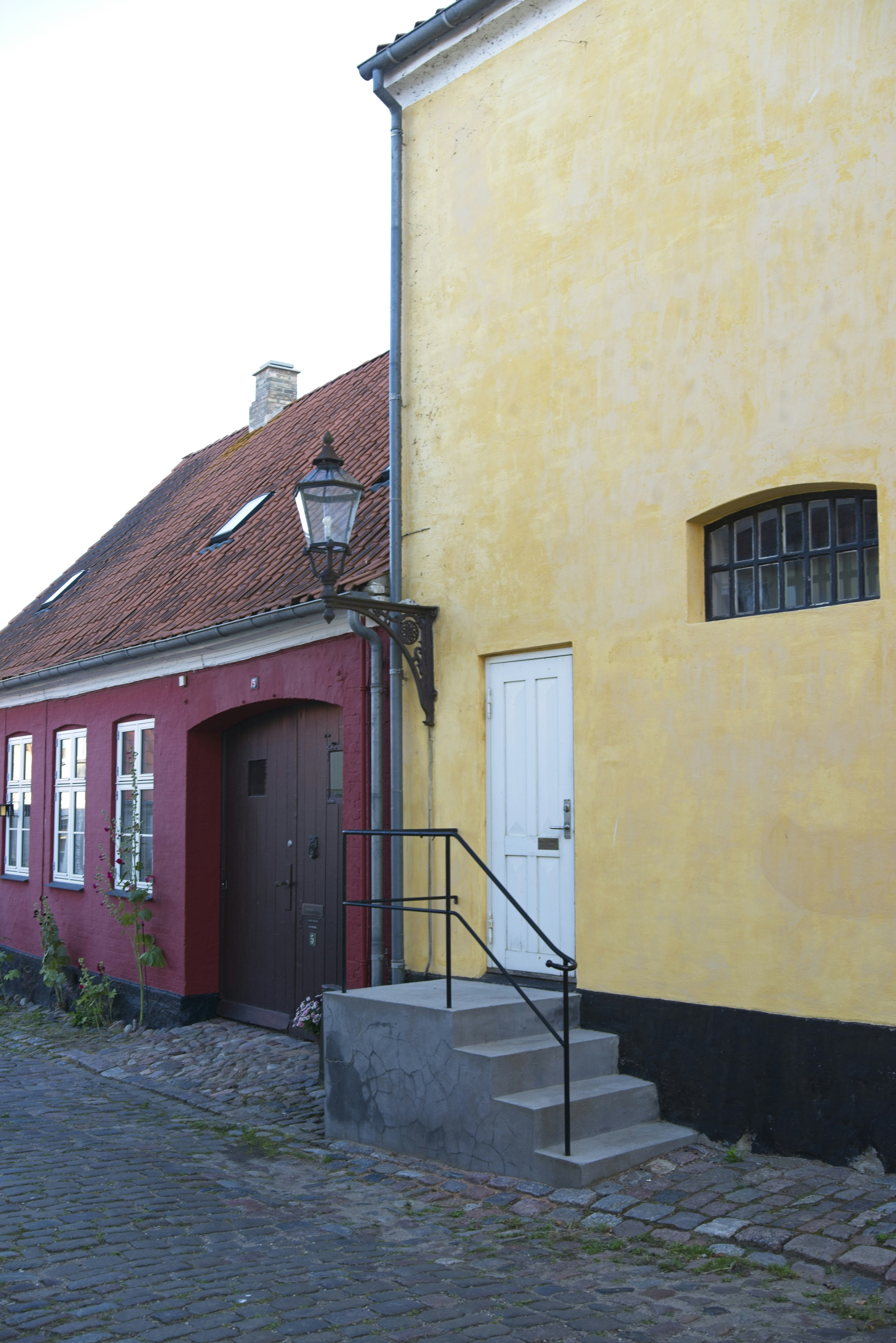 A deep red house with a heavy wooden door sits beside a tall yellow building with a simple white entrance, their contrasting facades meeting along a quiet cobblestone street in Præstø. An old-fashioned street lamp mounted on an ornate bracket adds character to the corner, while stone steps and a black railing lead up to the yellow building's door. Hollyhocks bloom modestly against the red wall. The muted evening light and understated architecture reflect the quiet, everyday charm of a small Danish town in late summer.