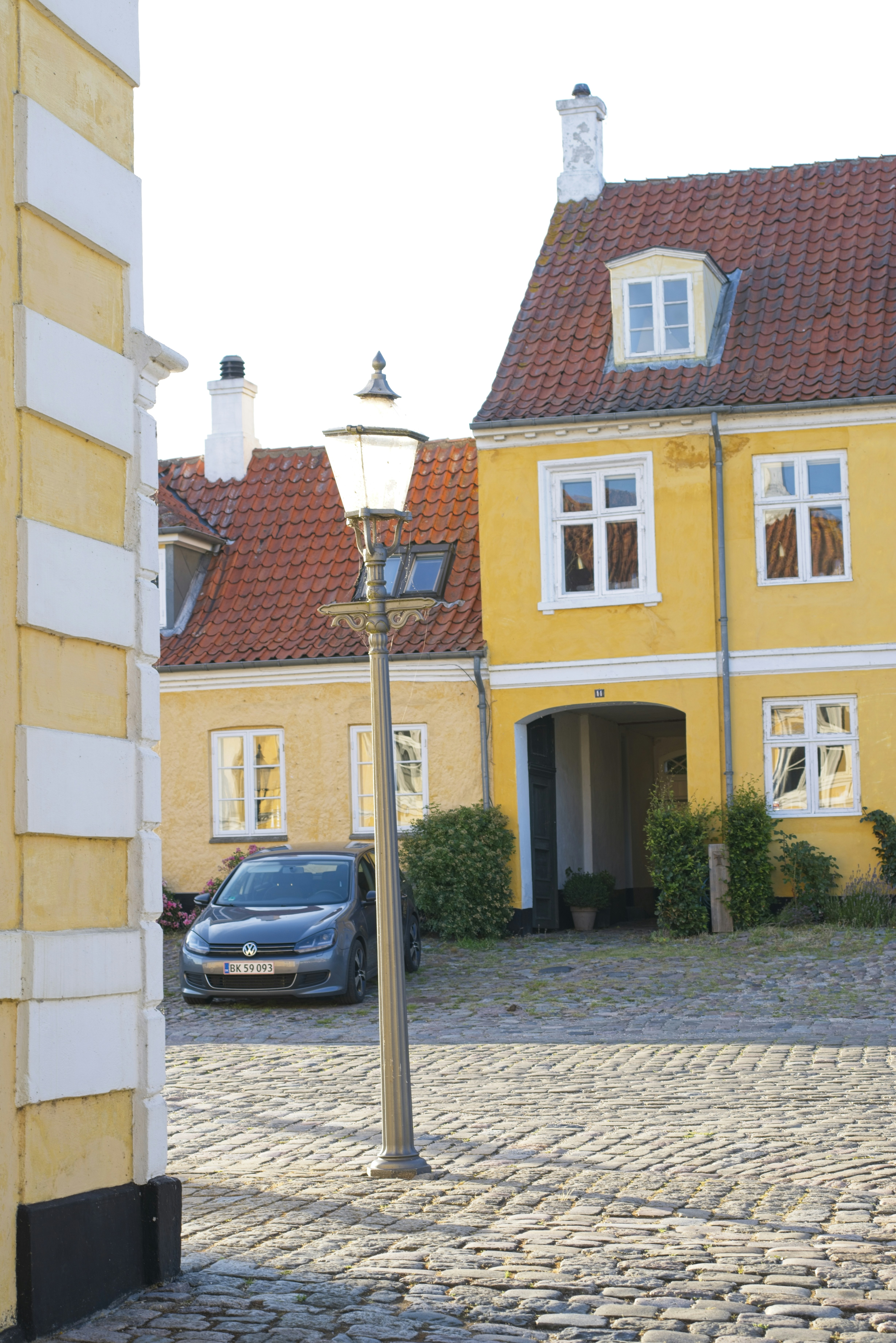 A tall lantern-style street lamp stands sentinel in a quiet cobblestone courtyard in Præstø, framed by rows of yellow buildings with red-tiled roofs and white-trimmed windows. A gray Volkswagen is parked before an open archway leading into a shaded passage marked "11," while low green shrubs soften the building's base. Striped yellow-and-white pilasters on the corner building add subtle architectural rhythm to the scene. The soft, hazy evening light and warm building tones capture the timeless, unhurried atmosphere of Præstø's historic streets in July.