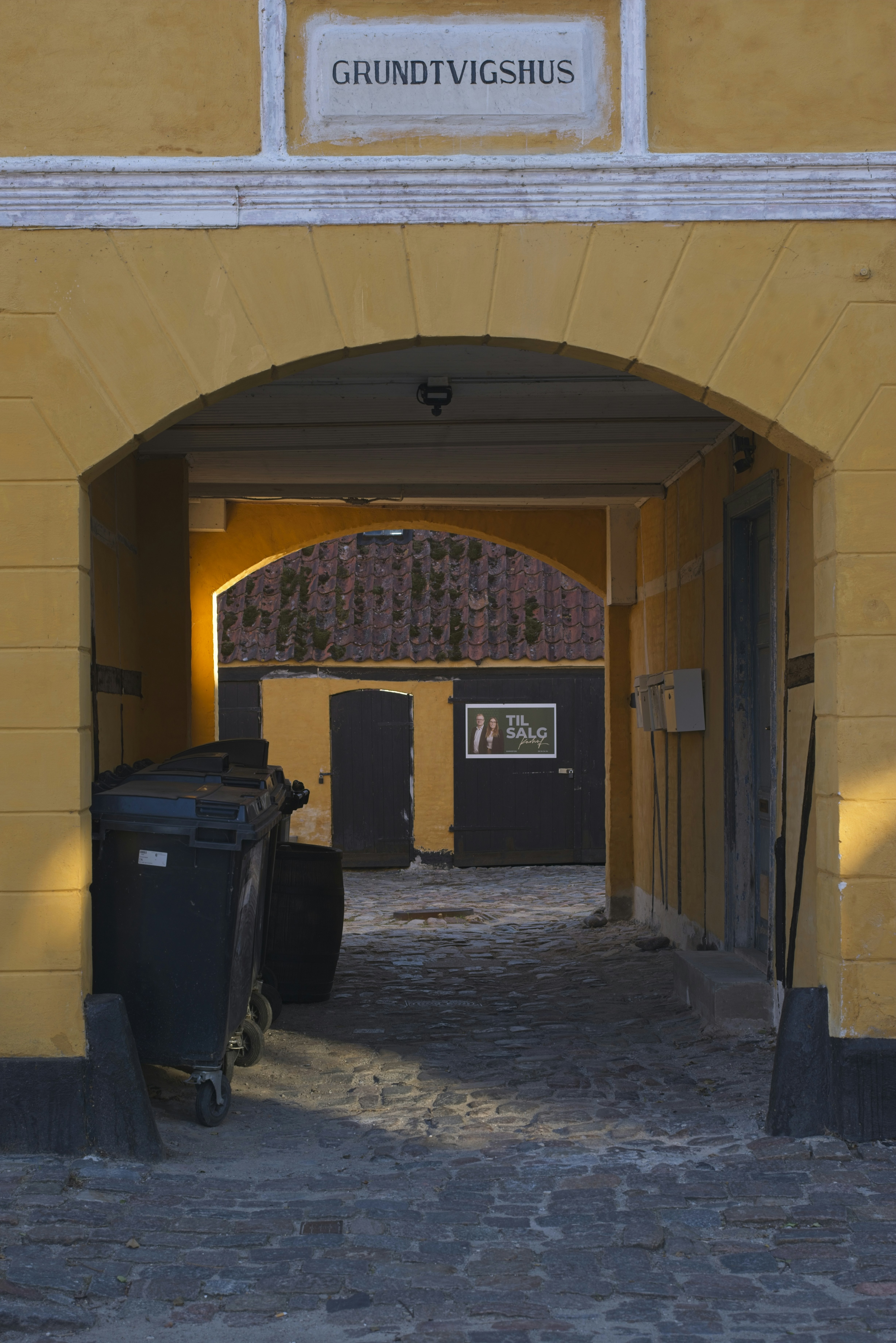 A yellow archway marked "Grundtvigshus" leads into a shadowed cobblestone passage in Præstø, revealing weathered dark doors and a moss-covered tiled roof beyond. The Grundtvig House (Danish: Grundtvigshus) is a complex of historic buildings at Torvestræde 7 in Præstø, Vordingborg Municipality, Denmark. It takes its name after N. F. S. Grundtvig, who lived there for a year and a half in the early 1820s.