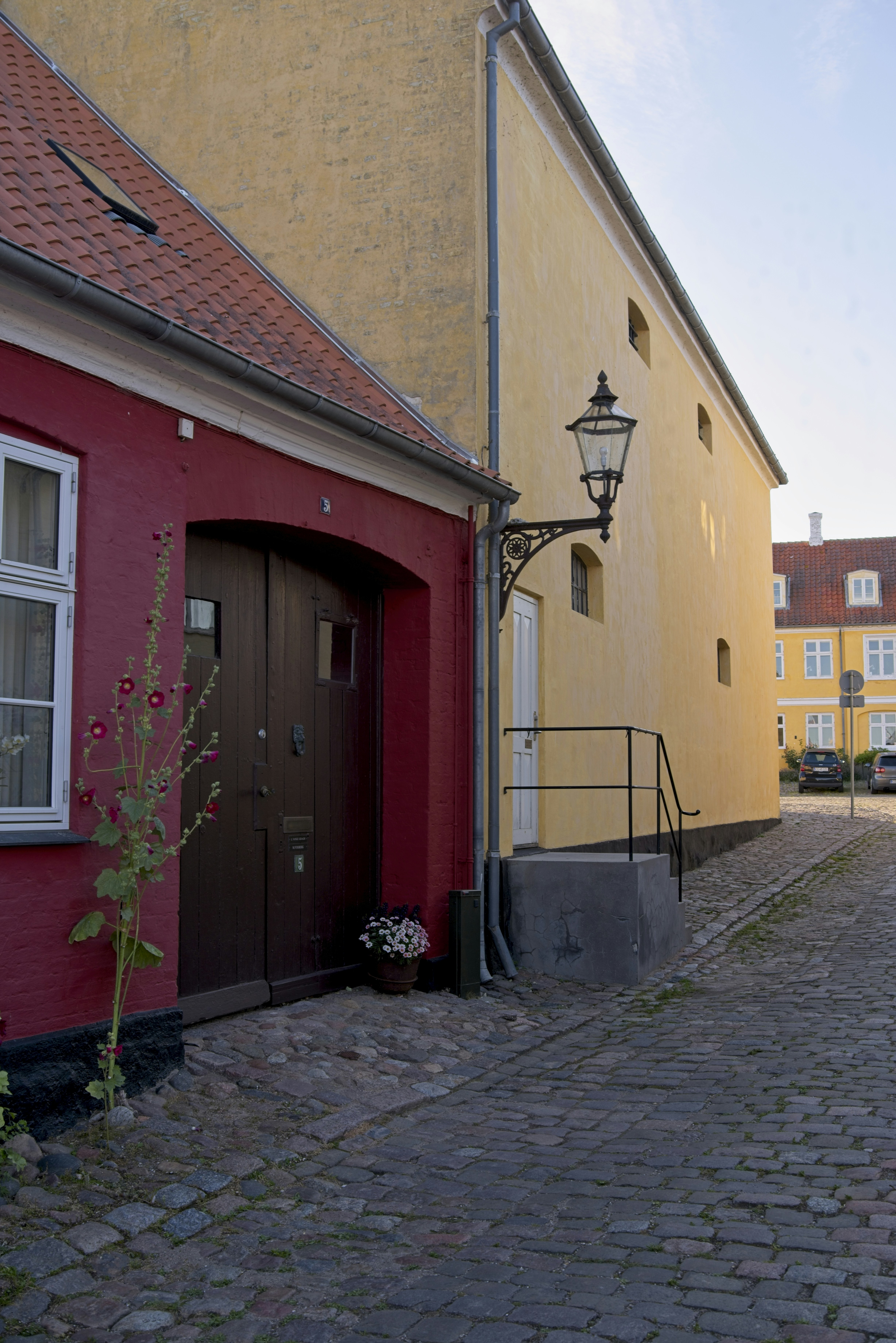 A striking contrast of colors unfolds along a quiet cobblestone lane in Præstø, where a deep red house with a heavy wooden door and blooming hollyhocks meets a tall pale-yellow wall crowned by an ornate street lamp. Small arched windows and a modest white door punctuate the yellow facade, leading toward more traditional houses further down the street. The soft evening light enhances the warm hues of both buildings. This quiet corner captures the layered architectural charm of Præstø's historic streets on a hot July evening.