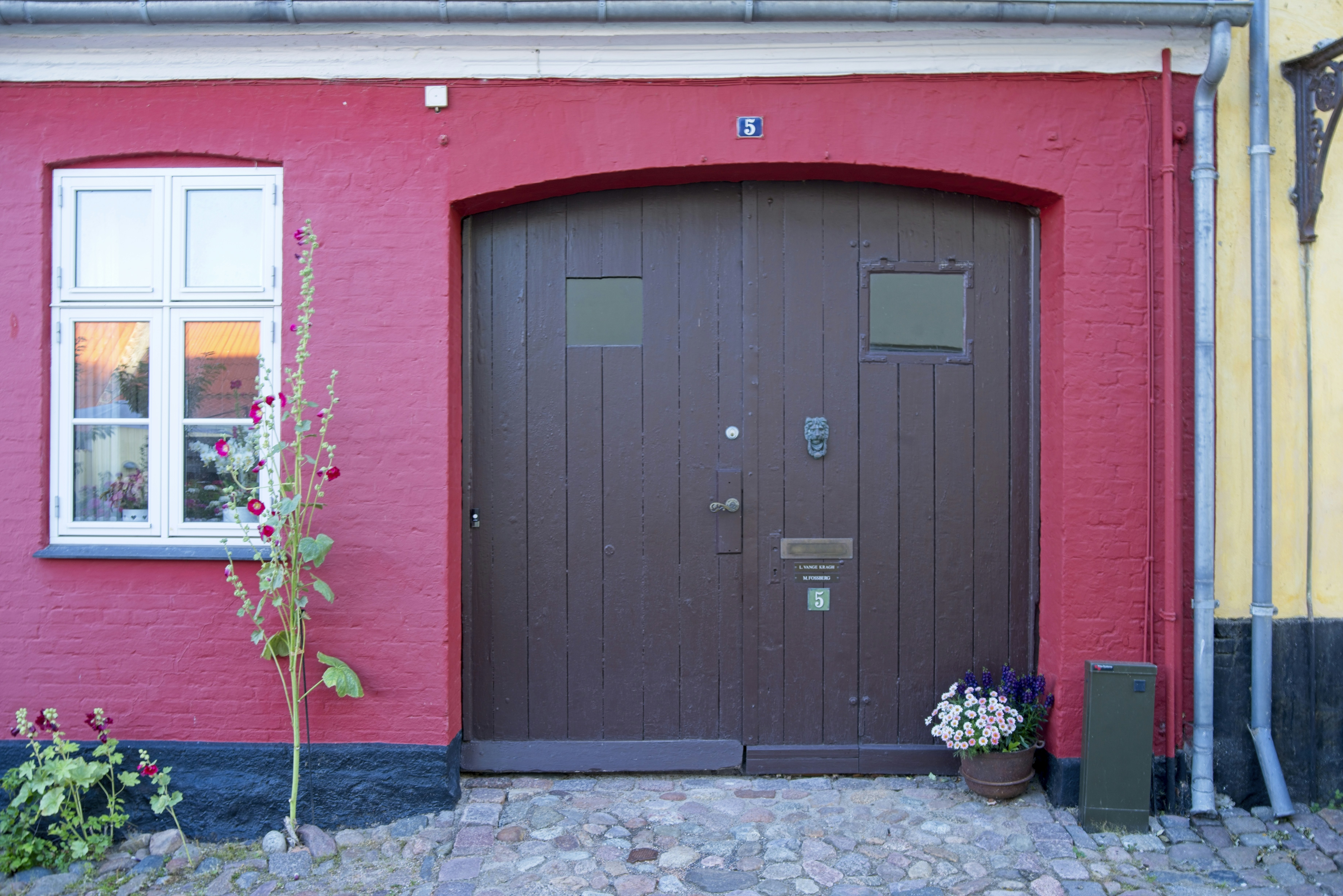 A weathered dark-brown double door with a lion-head knocker stands framed by a deep pink facade at house number 5 in Præstø, its arched entrance flanked by a tall hollyhock and a white-framed window. Potted flowers add soft color near the cobblestone entryway, while a nameplate reveals residents' names beside the mail slot. The rich pink and brown tones create a striking contrast against the warm evening light. This detailed doorway captures a quintessential slice of Præstø's colorful, historic streetscape on a hot July evening.