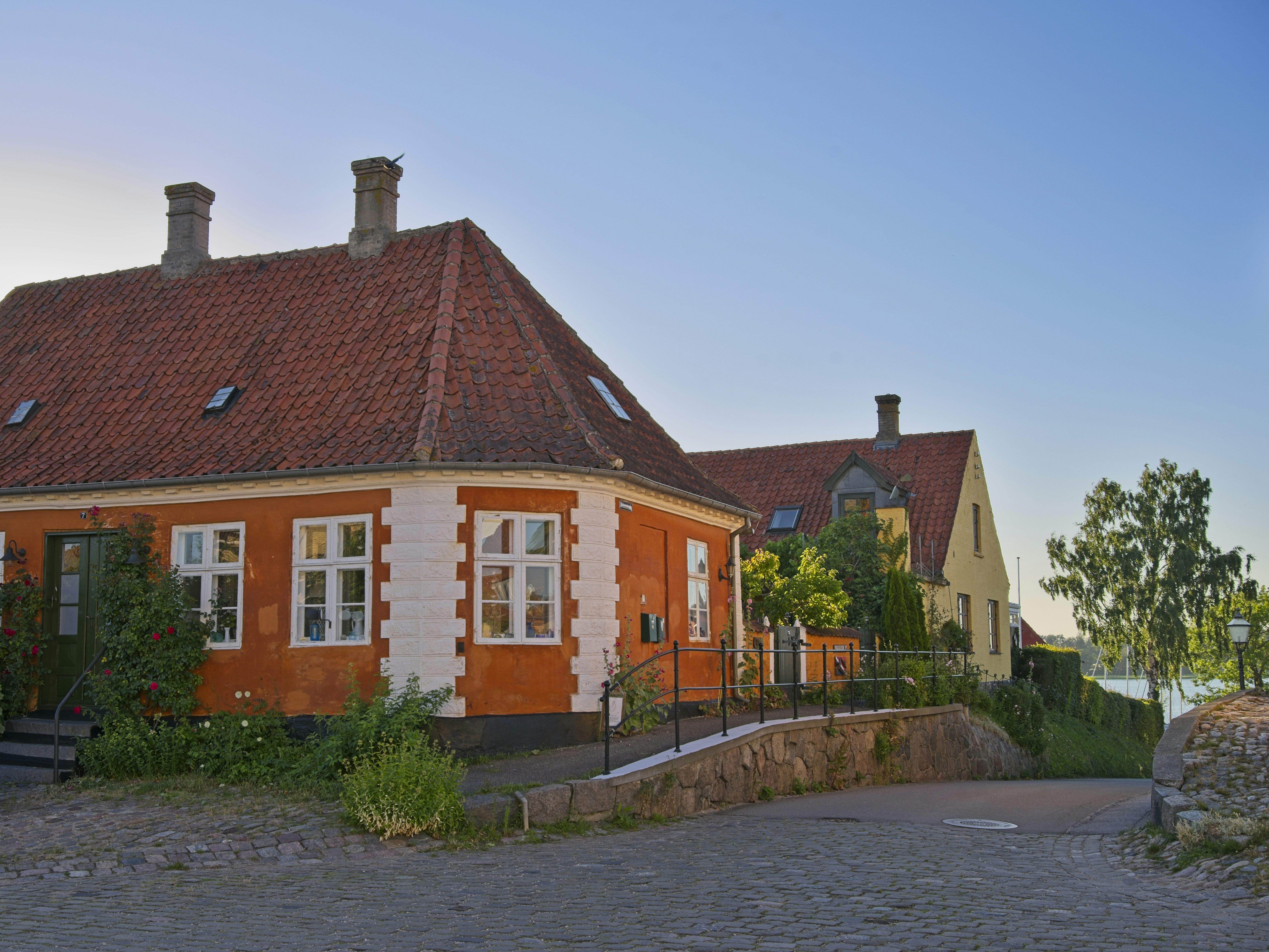 A charming orange corner house with crisp white quoins and white-framed windows anchors this quiet cobblestone corner near Præstø's waterfront, its rounded facade softened by climbing roses and wildflowers. A stone retaining wall and iron railing curve toward a glimpse of the harbor visible in the distance. Neighboring rooftops in red tile and mustard-yellow walls complete the picturesque streetscape. The warm evening light and coastal proximity capture the relaxed, sun-soaked character of Præstø on a hot July evening.