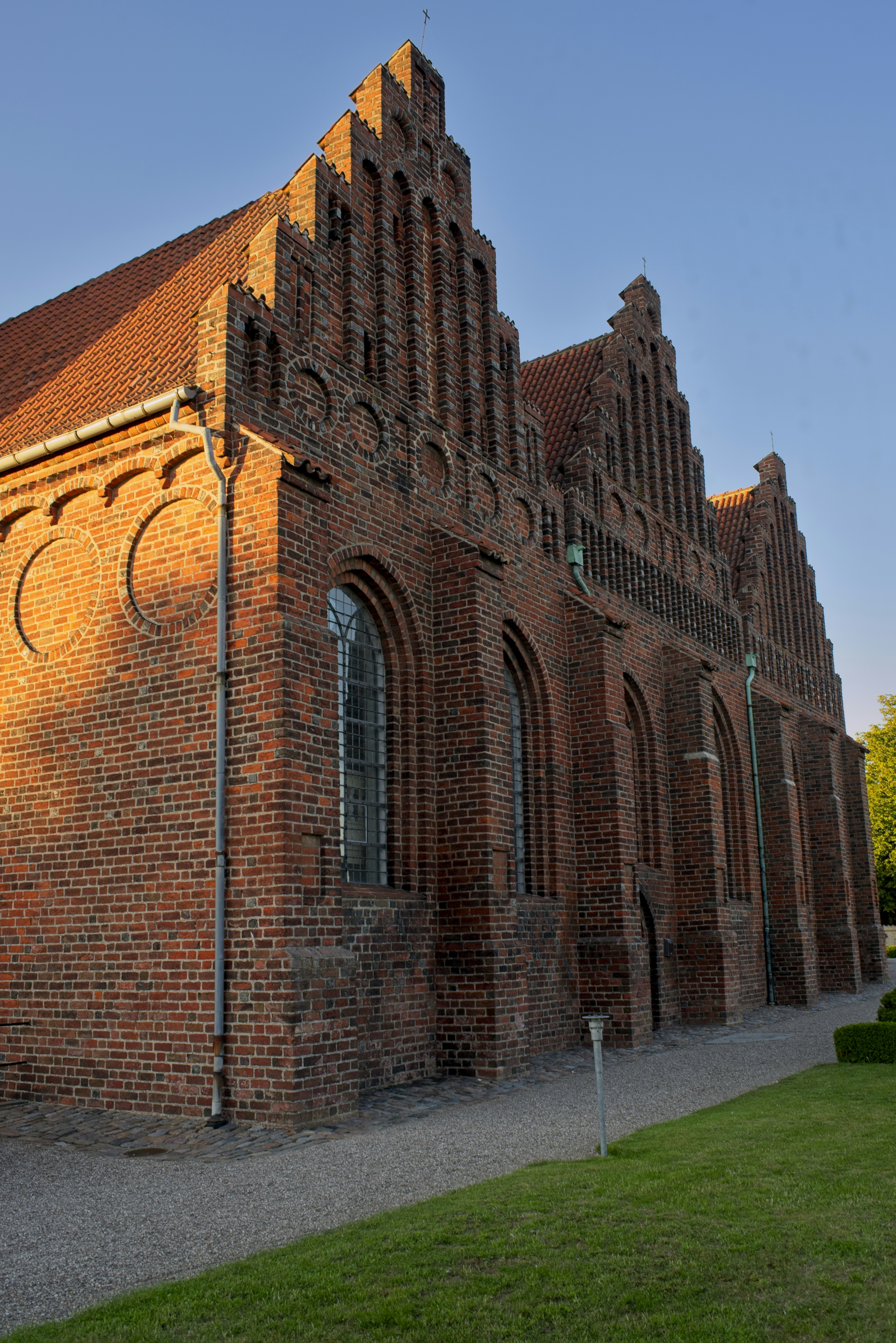 The intricate stepped brick gables of Præstø Church catch the golden light of a hot July evening, their decorative circular motifs and pointed arched windows showcasing fine medieval Gothic craftsmanship. Tall, narrow finials rise dramatically against a clear blue sky, while a gravel path and neatly trimmed lawn lead along the church's red-brick facade. The warm evening glow accentuates the deep textures of the aged brickwork, highlighting centuries of history. This striking architectural view captures the enduring grandeur of Præstø's historic church at sunset.