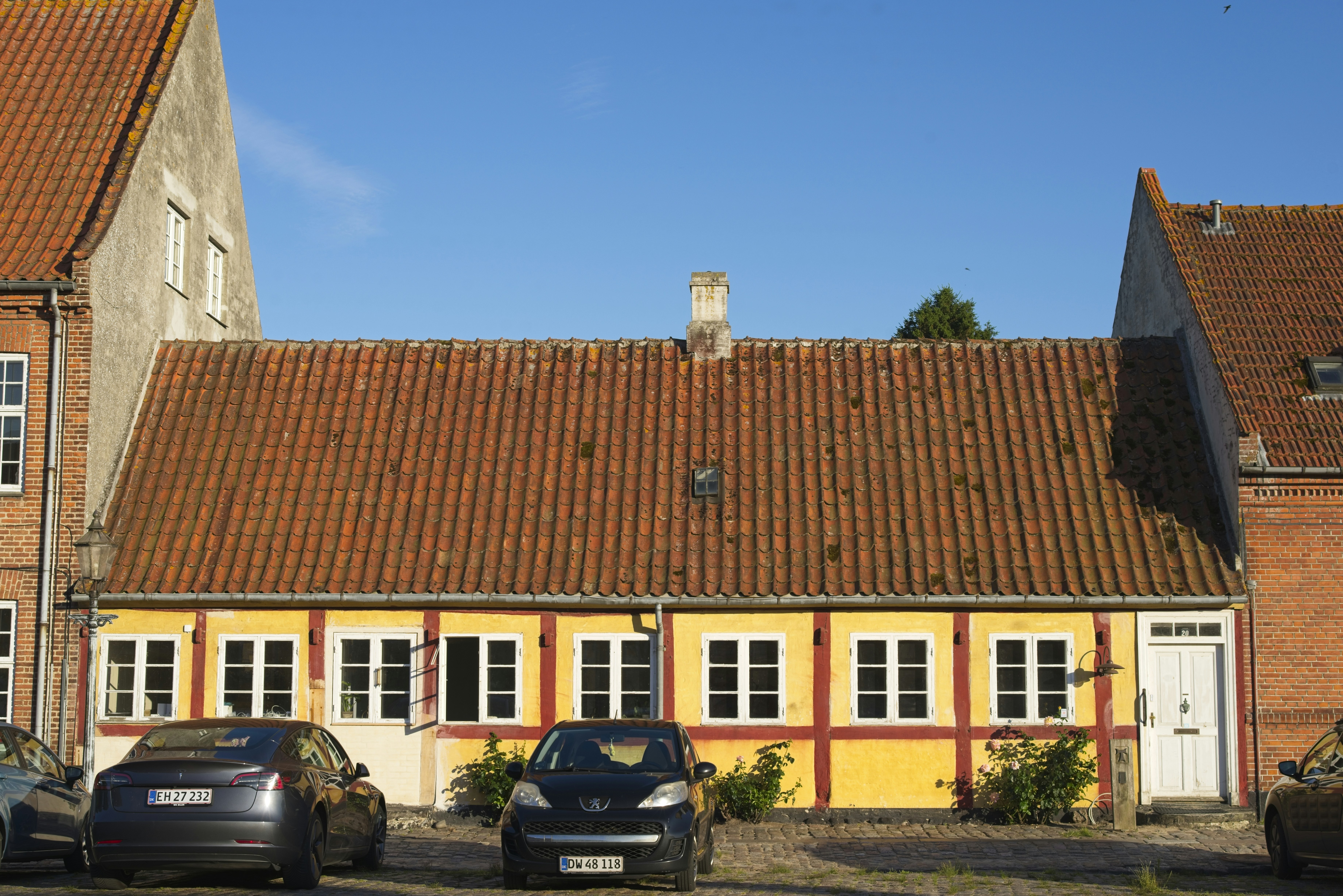 A charming half-timbered house with yellow and red trim stretches along a quiet street in Præstø, its steep tiled roof and white-framed windows glowing under a clear blue sky. Parked cars line the cobblestone street out front, while a taller brick building rises beside it, adding contrast to the streetscape. Small rose bushes soften the base of the timber facade near the entrance. The warm evening light and traditional half-timbered construction capture a classic slice of Danish small-town charm on a hot July evening.