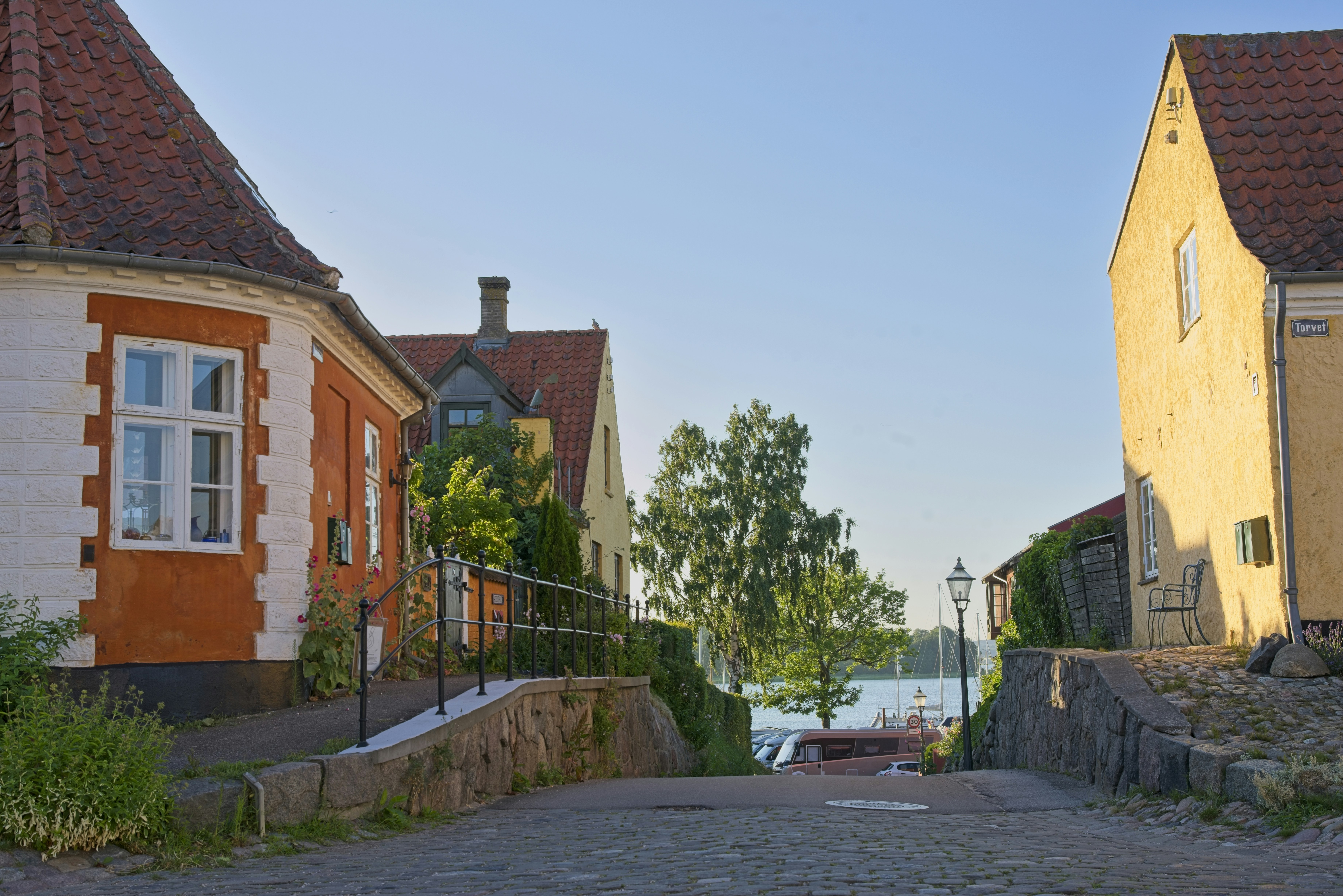 A gently sloping cobblestone street on Torvet in Præstø leads down toward glimpses of sailboats and calm water in the distance, framed by charming orange, yellow, and cream houses with red-tiled roofs. Wrought-iron railings and blooming hollyhocks line the path, while a leafy tree marks the transition from town to harbor. The warm evening sun bathes the facades in golden light, highlighting the town's picturesque descent to the waterfront. This scenic street captures the timeless charm of Præstø's harbor town character on a hot July evening.