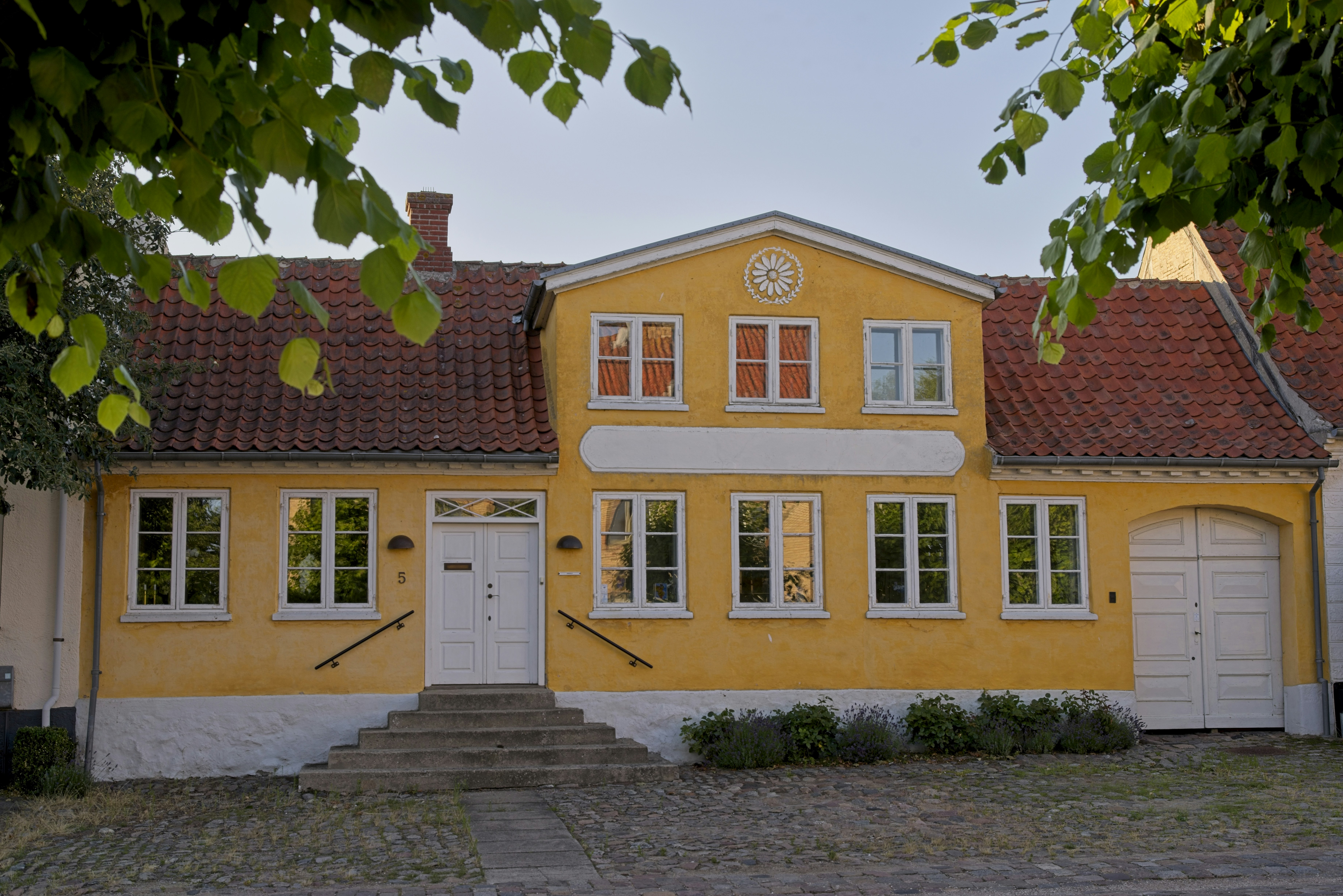 A grand yellow townhouse at number 5 stands framed by overhanging green leaves, its symmetrical white-trimmed windows and ornate gable rosette catching the soft evening light. A stone staircase leads up to double white doors, flanked by wrought-iron handrails and low garden shrubs. The red-tiled roof and tall brick chimney add warmth to the classic Danish facade, while a rounded white archway marks a garage door to the right. This elegant townhouse captures the timeless charm of Præstø's historic streets on a hot July evening.