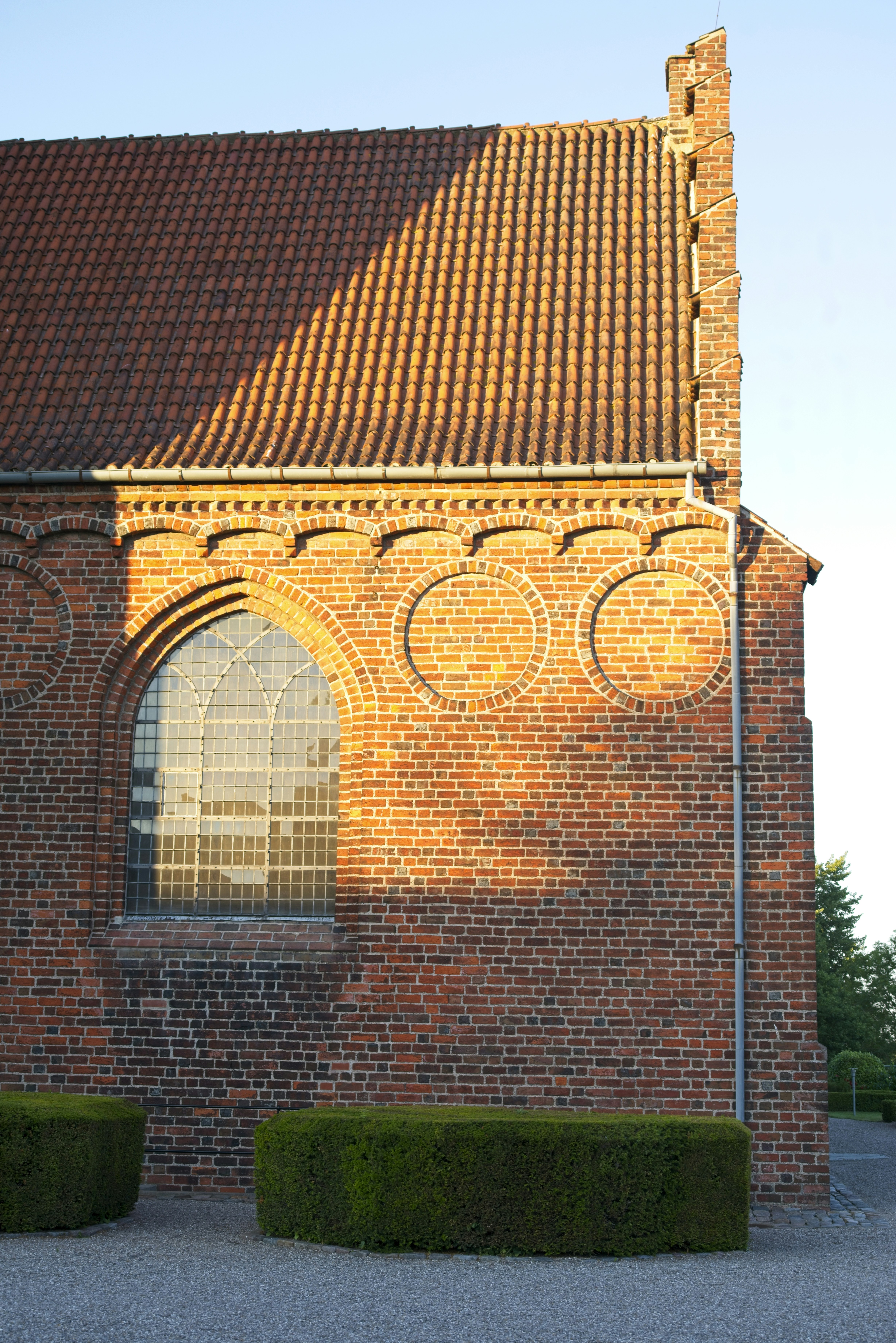 The medieval brick facade of Præstø Church glows richly in the evening sun, its decorative circular motifs and tall arched window catching warm golden light against a deep blue sky. A stepped brick gable and red-tiled roof rise sharply above neatly trimmed hedges bordering the churchyard. The rich texture of the aged brickwork and precise architectural detailing highlight centuries of craftsmanship. This close-up view captures the timeless beauty of Præstø's historic church bathed in the warm glow of a hot July evening.