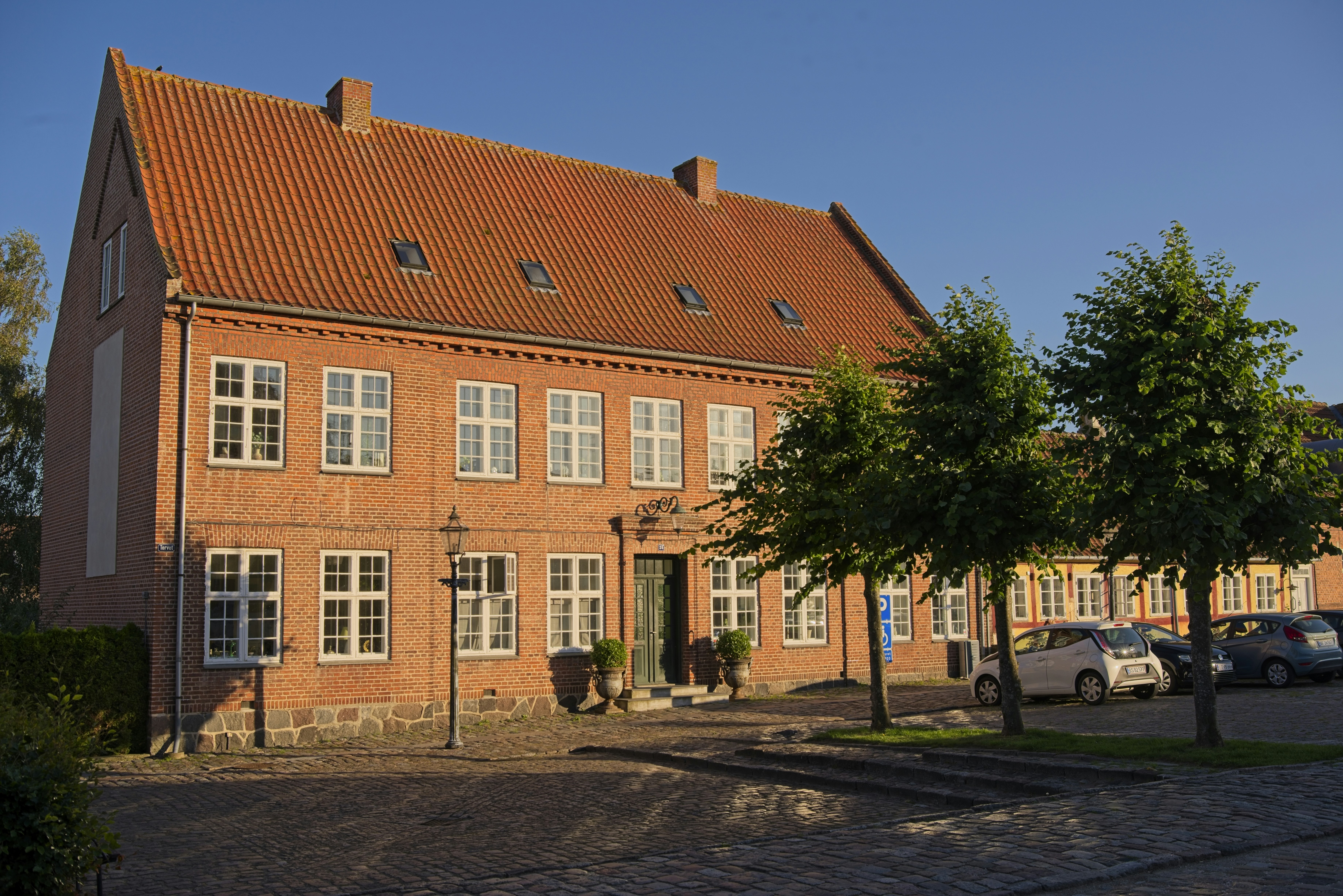 A stately red-brick building on Torvet stretches along a cobblestone square in Præstø, its steep tiled roof punctuated by small dormer windows and tall brick chimneys. Neat rows of white-framed windows and a dark green entrance door with potted topiaries add classic detail to the facade. Leafy trees and a scattering of parked cars line the street in the warm evening light. The long shadows and golden glow on the brickwork capture the quiet, sun-soaked stillness of Præstø's historic town square on a hot July evening.