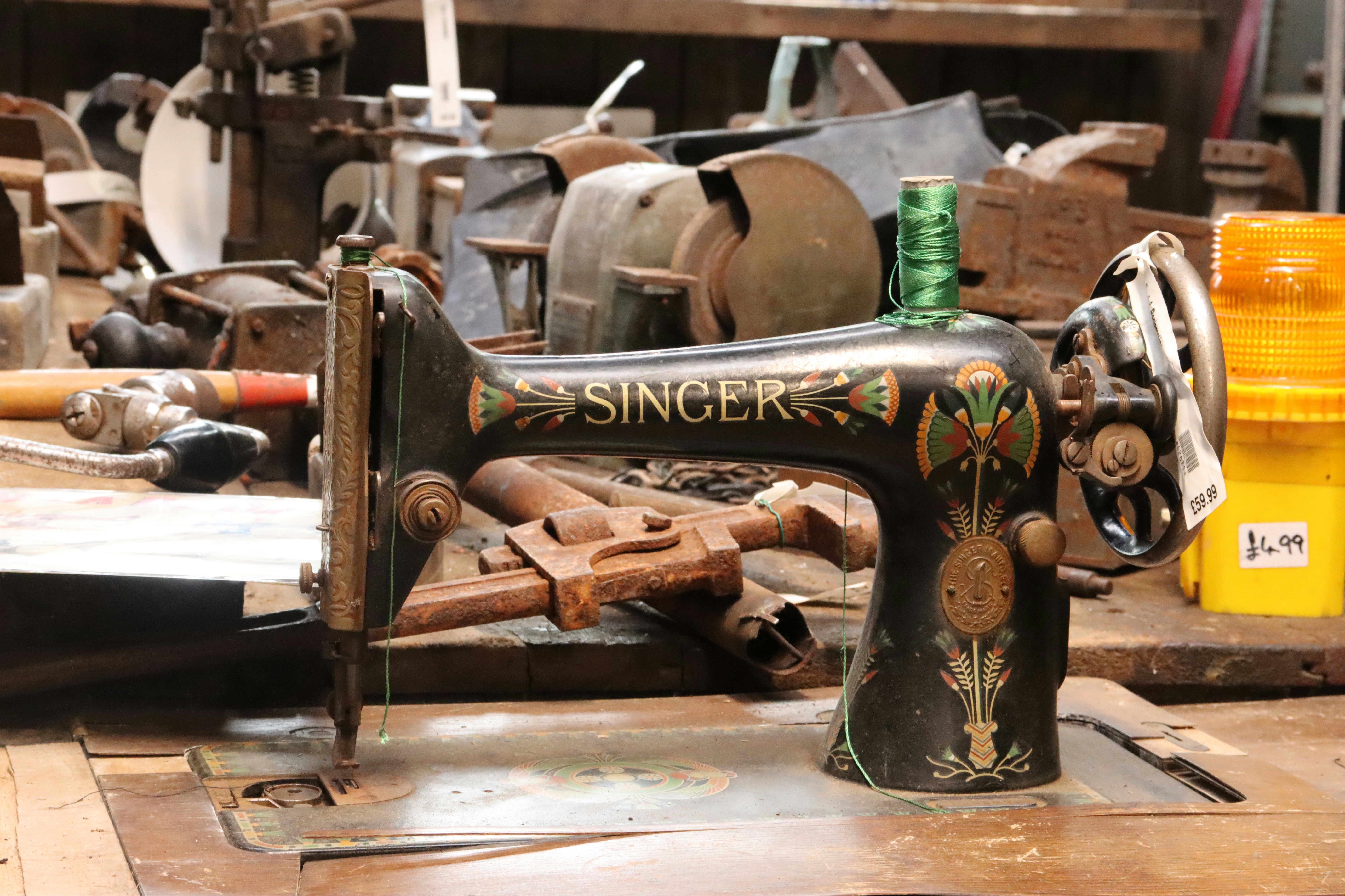 A vintage Singer sewing machine sits in a cluttered workshop, its ornate floral detailing faded by time and dust, surrounded by assorted old tools and hardware.