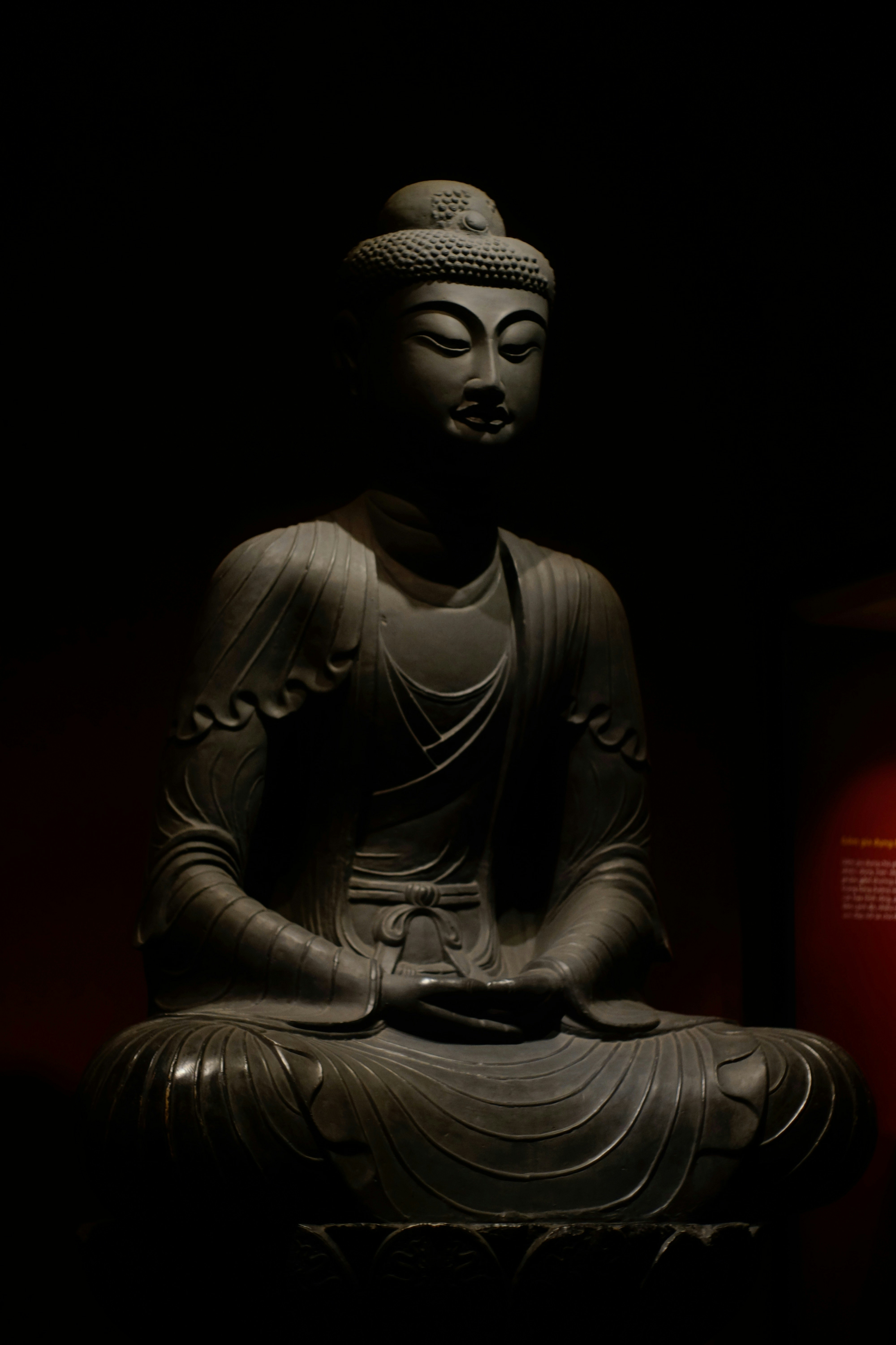 Stone Buddha statue illuminated in a dark gallery, subtle highlights revealing serene facial features and drapery details.