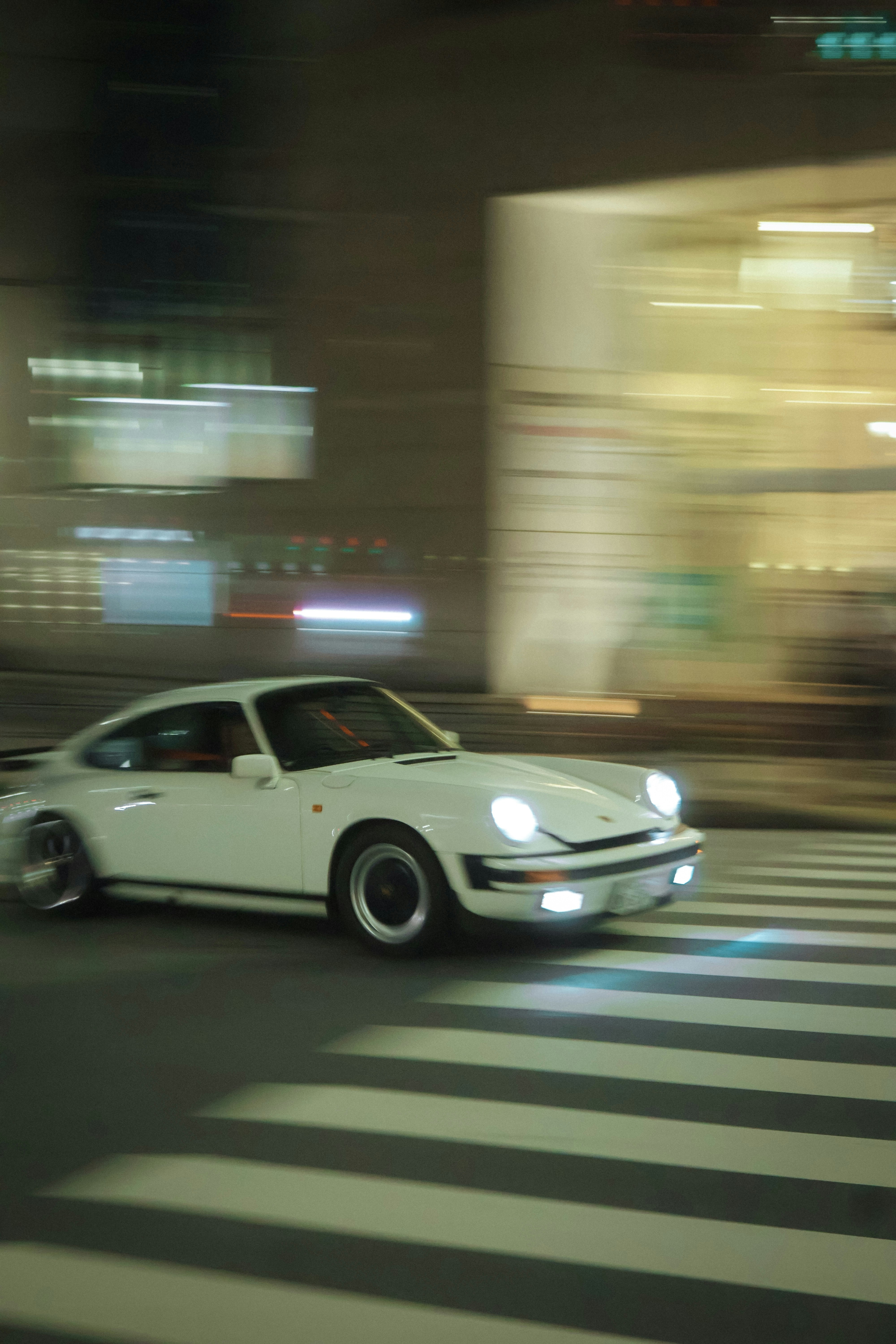Nighttime street scene shows a white Porsche speeding across a crosswalk with motion blur and city lights streaking in the background.