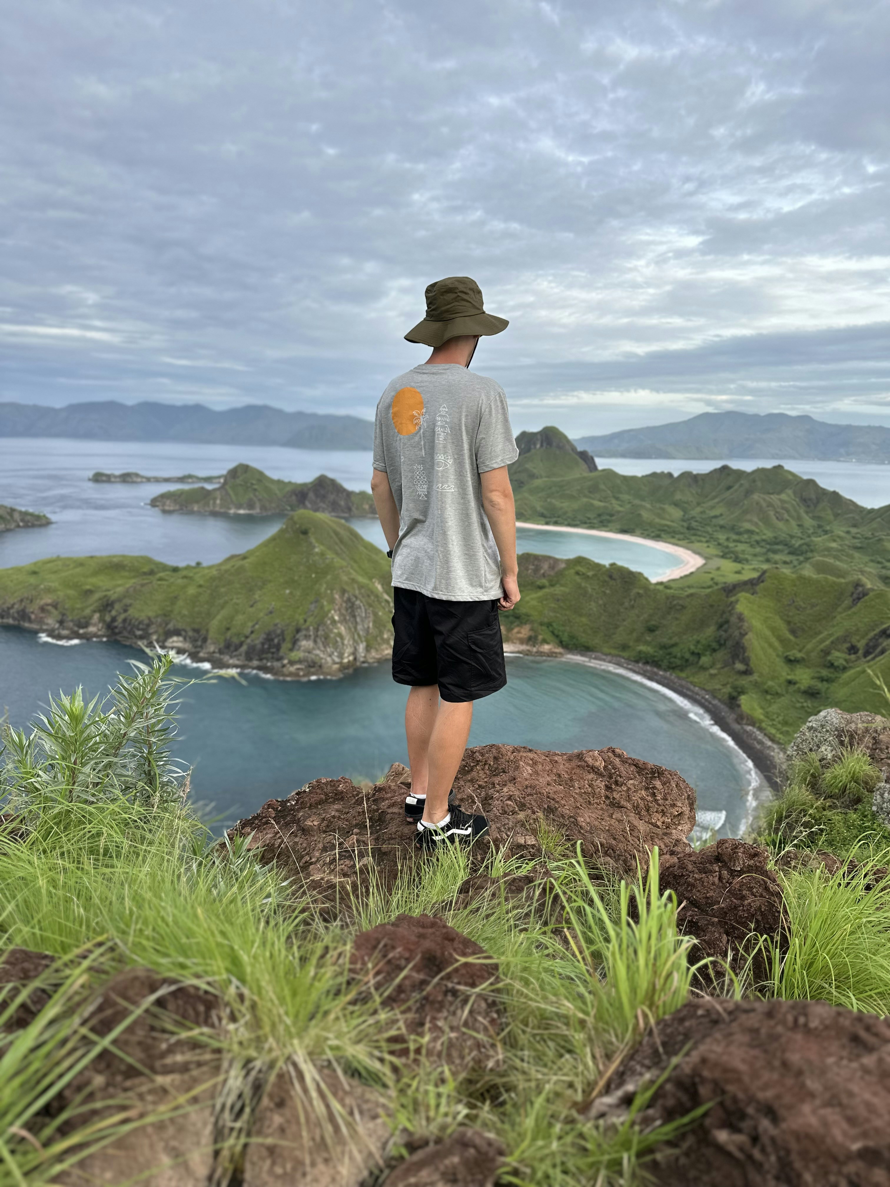 Man standing on a rocky overlook, facing a crescent bay and layered islands with lush green hills under a cloudy sky.