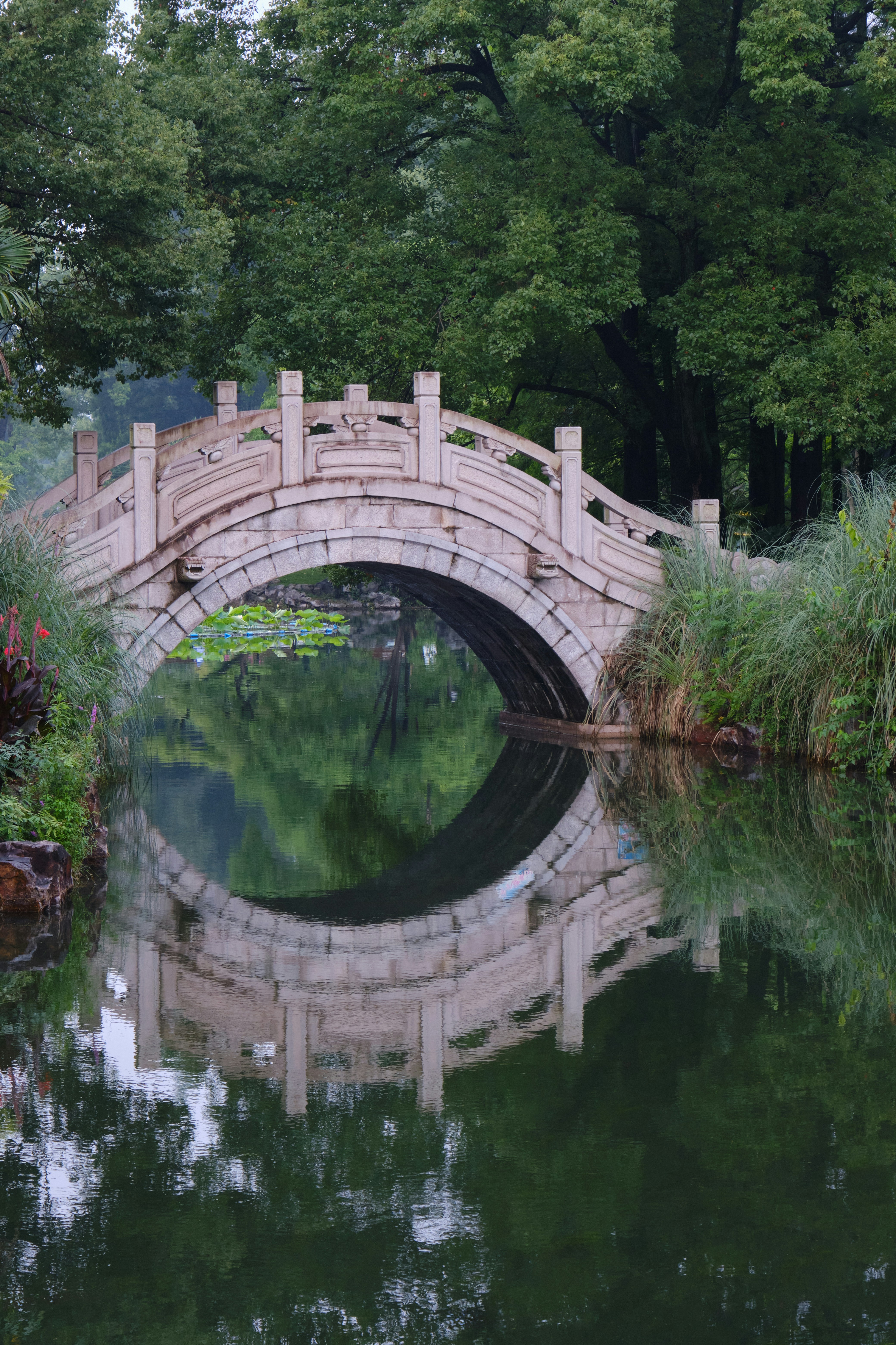 Stone arch bridge over a calm pond with lush greenery and a near-perfect reflection, creating a serene park scene.