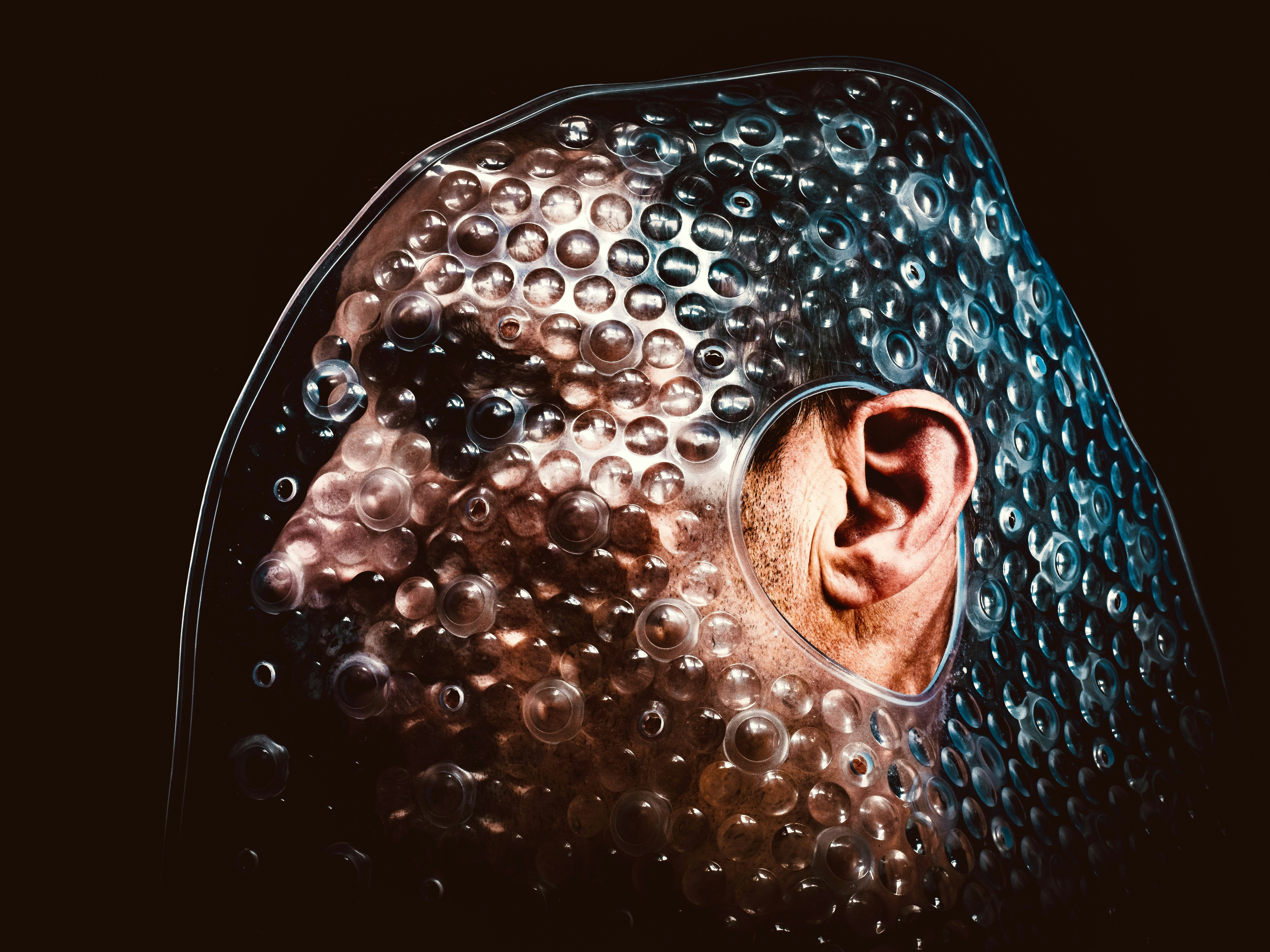 A close-up portrait of a person's head wrapped in a translucent bubble mask, revealing facial features through a pattern of circular bubbles. This is a staged, sculptural portrait emphasizing texture and atmosphere.