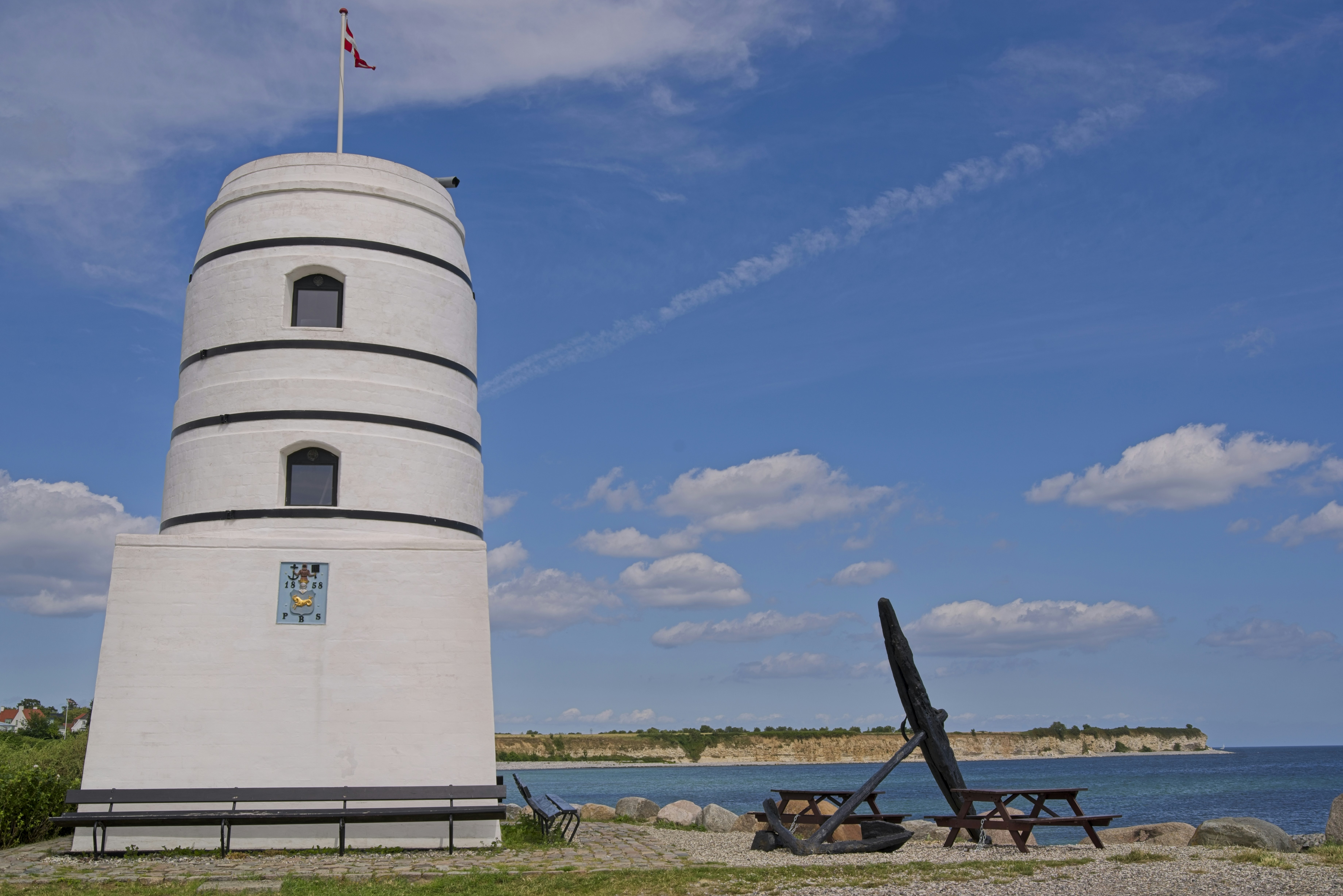 The historic Rødvig Flint Kiln stands against a clear blue sky in Rødvig, Stevns, Denmark, its tall chimney topped with a Danish flag on a sunny July day. This 3.5-metre square building, now fitted with three stacked rooms serving as a local museum, includes an observation platform offering coastal views. Wooden benches line its base, while a weathered old anchor and rustic picnic tables sit nearby along the gravel shore, framed by the sea and distant chalk cliffs — a charming blend of industrial heritage and seaside scenery.