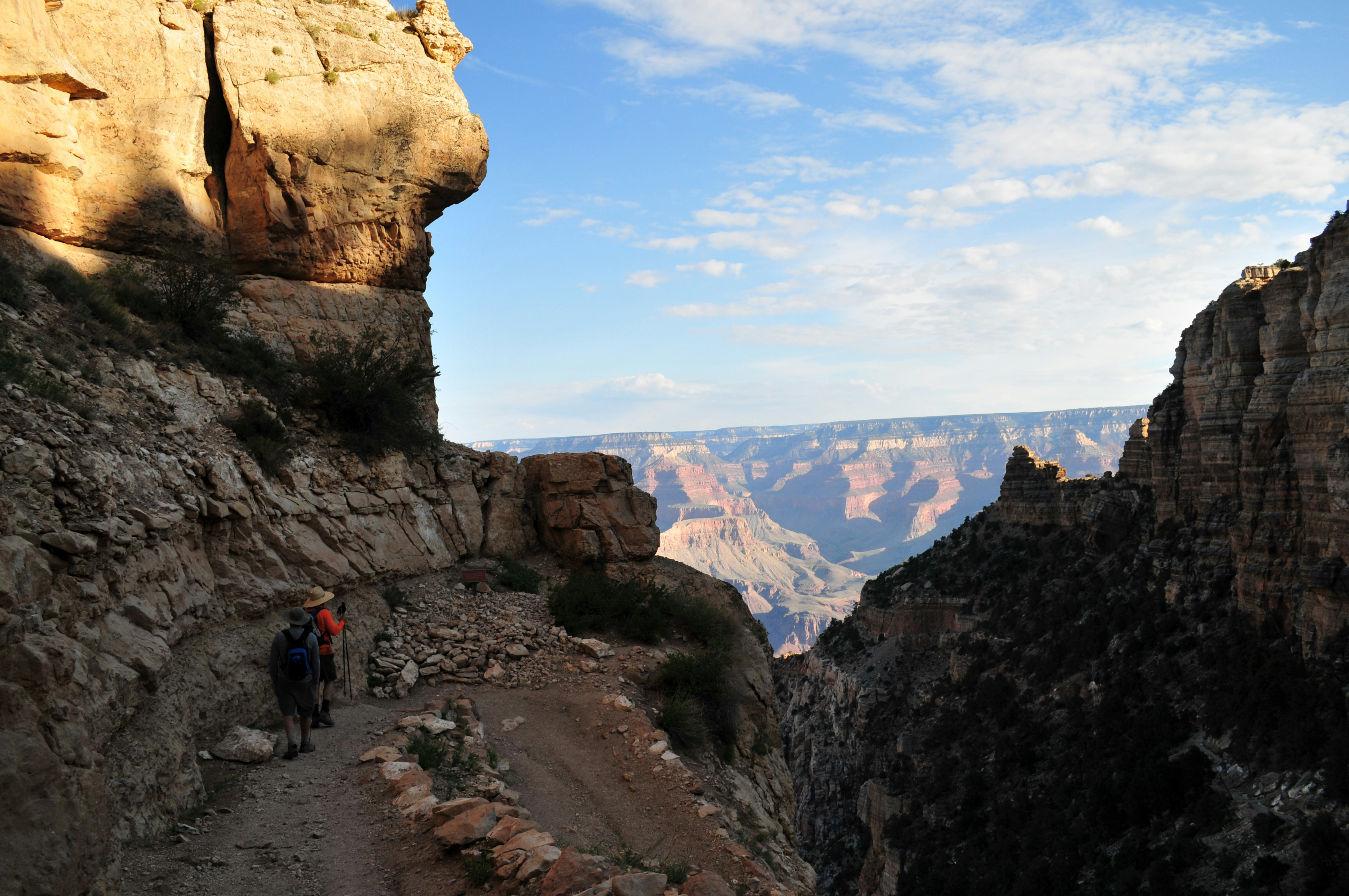 Hikers traverse a rugged trail along sunlit canyon walls with distant plateaus visible beyond a deep gorge.