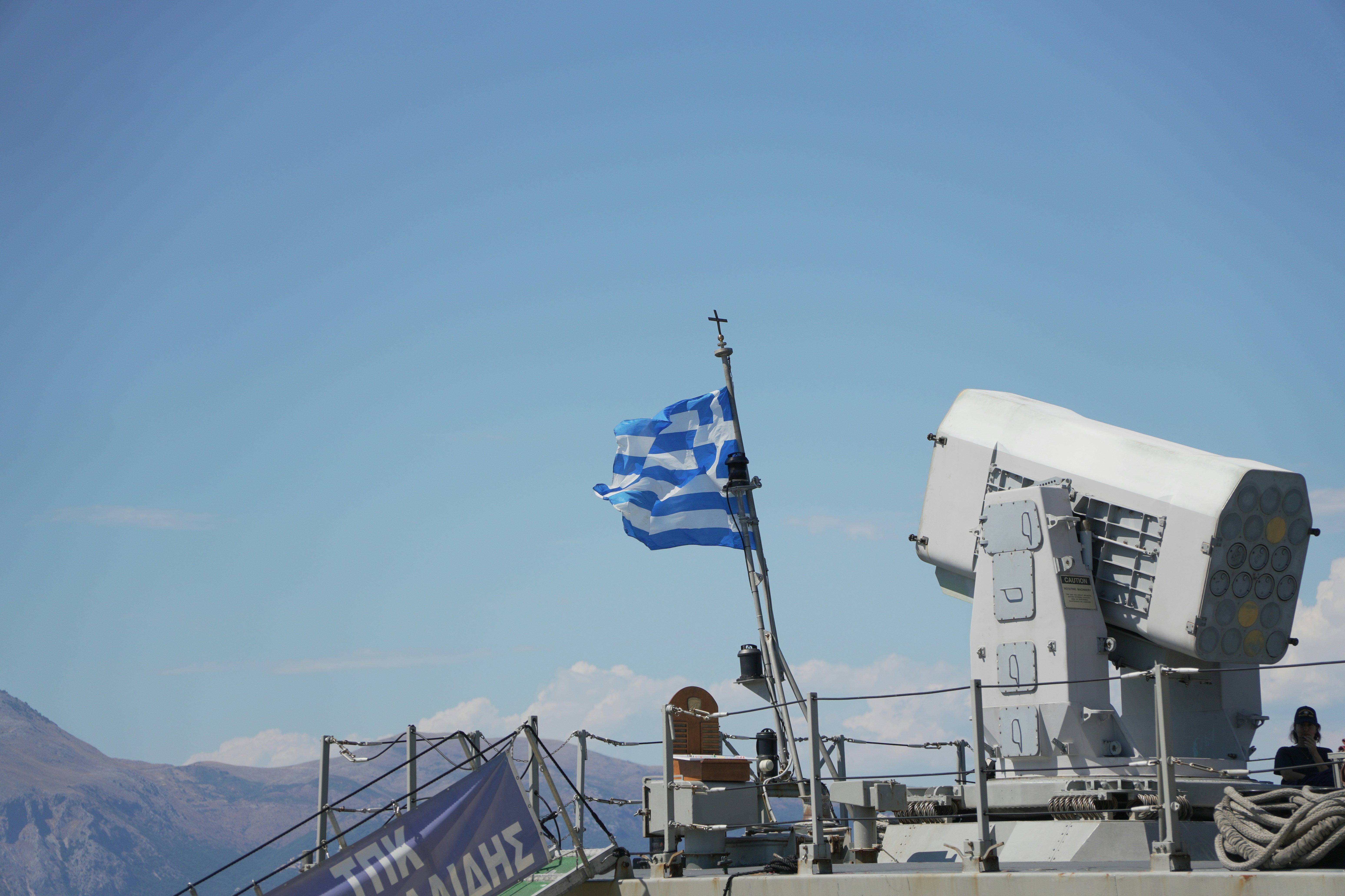 Greek Flag at the Deck Edge: Harbor Guns and Mountain Backdrop
