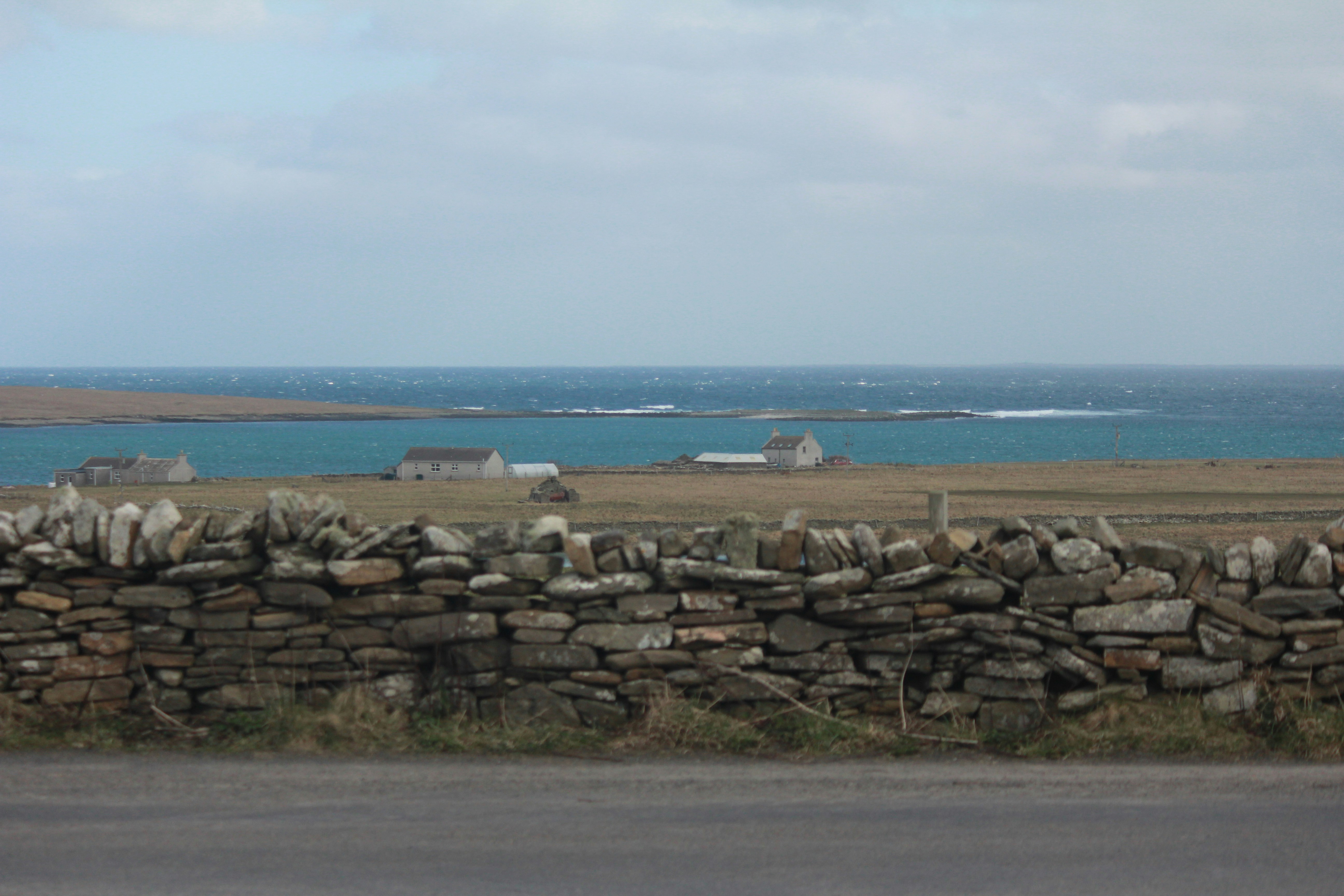 Low coastal field with a dry-stone wall, small cottages, and a blue sea on the horizon; a calm, rural shoreline scene with muted tones.