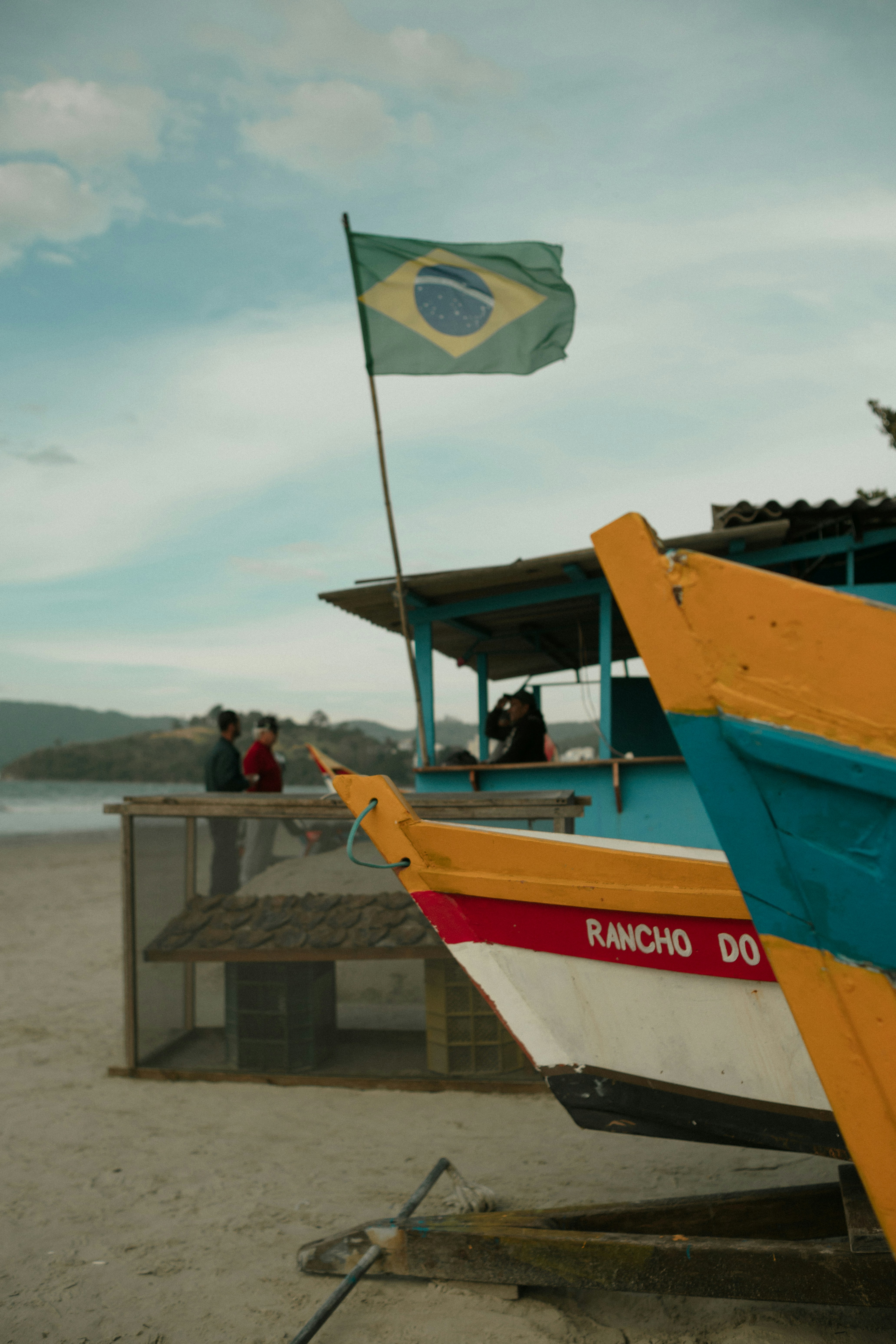 Brazilian flag flying over colorful boats on a sandy beach.