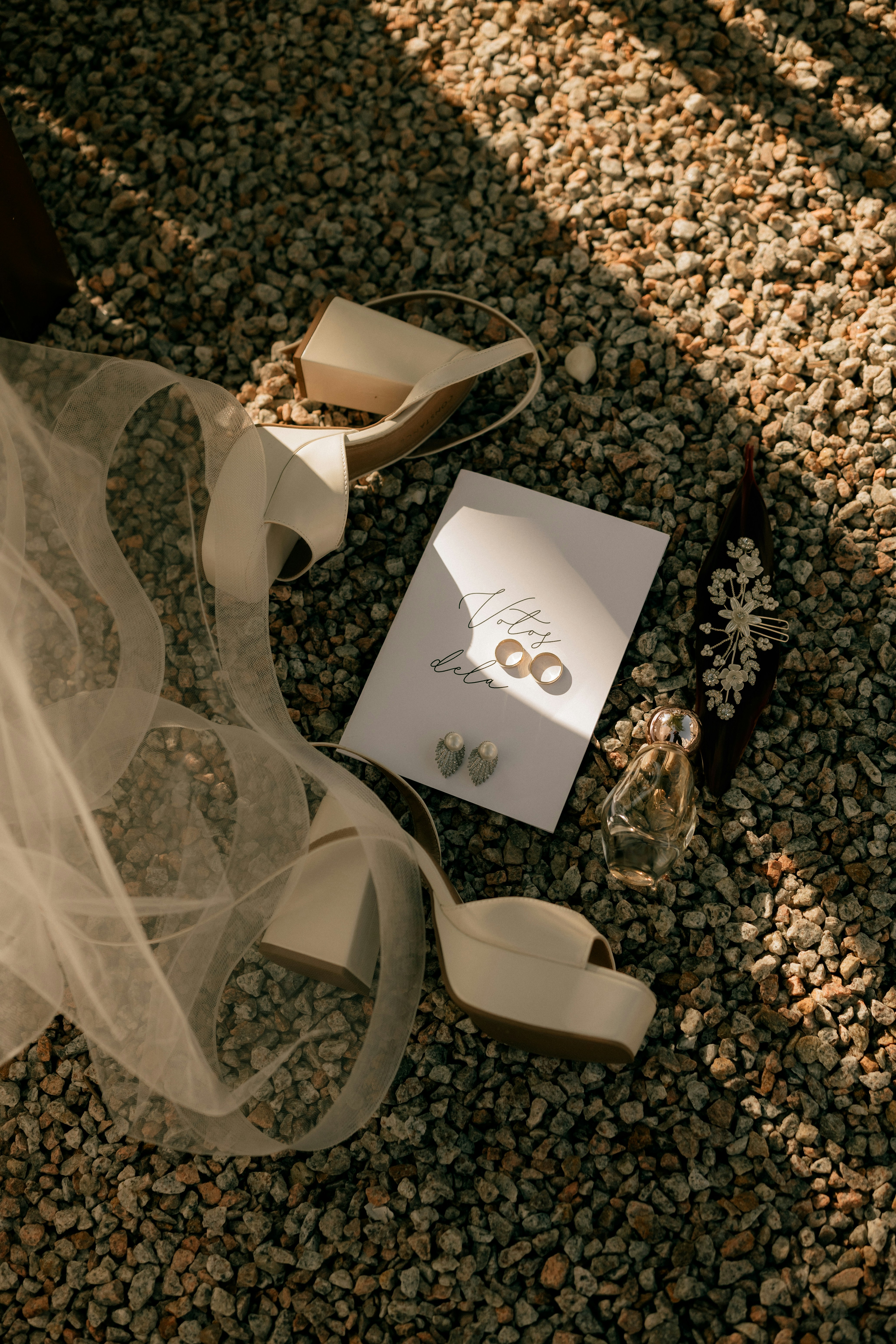 Elegant bridal details on gravel: heels, veil, and a small perfume bottle