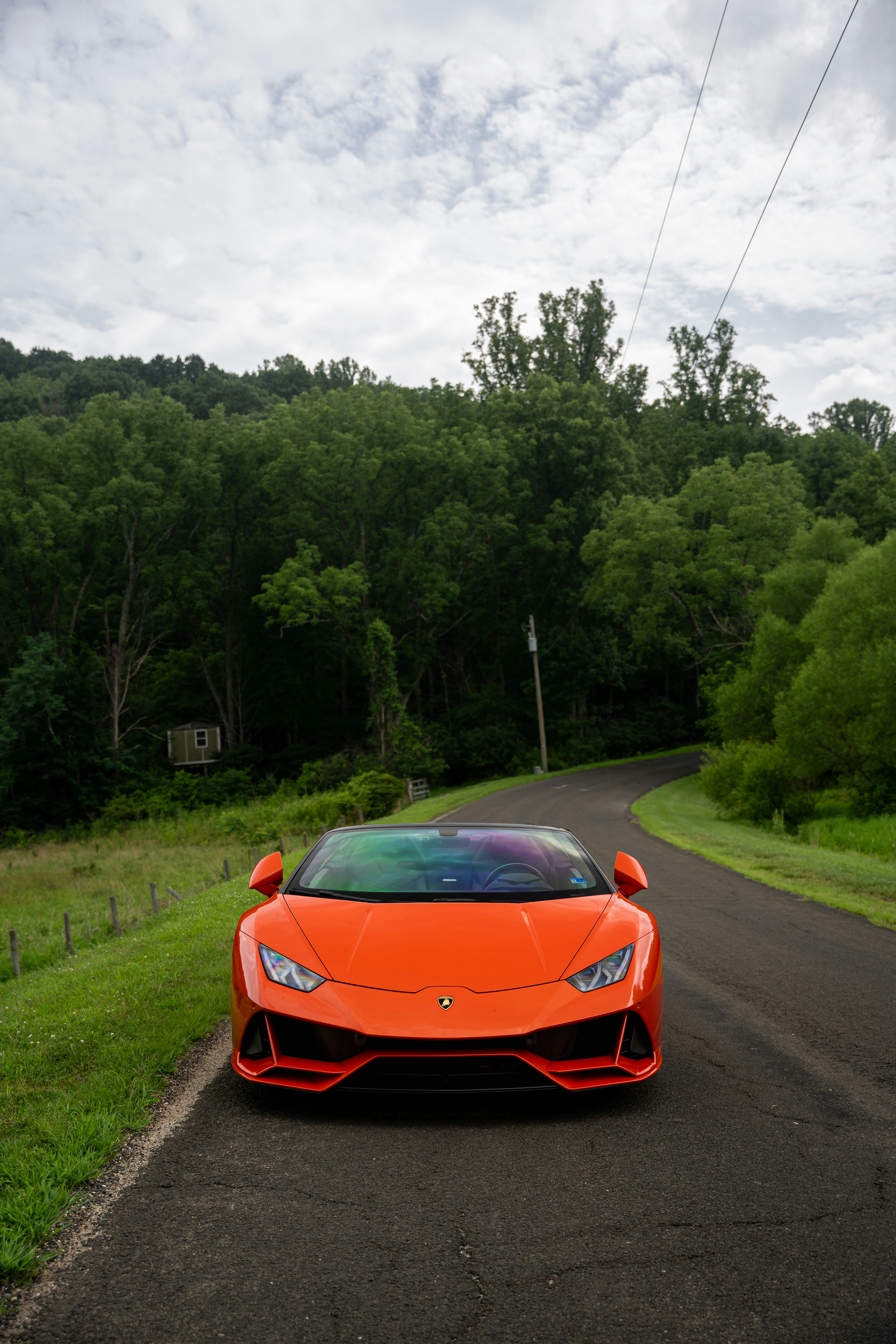 Orange Lamborghini parked on a winding rural road with dense green trees in the background, under a partly cloudy sky.