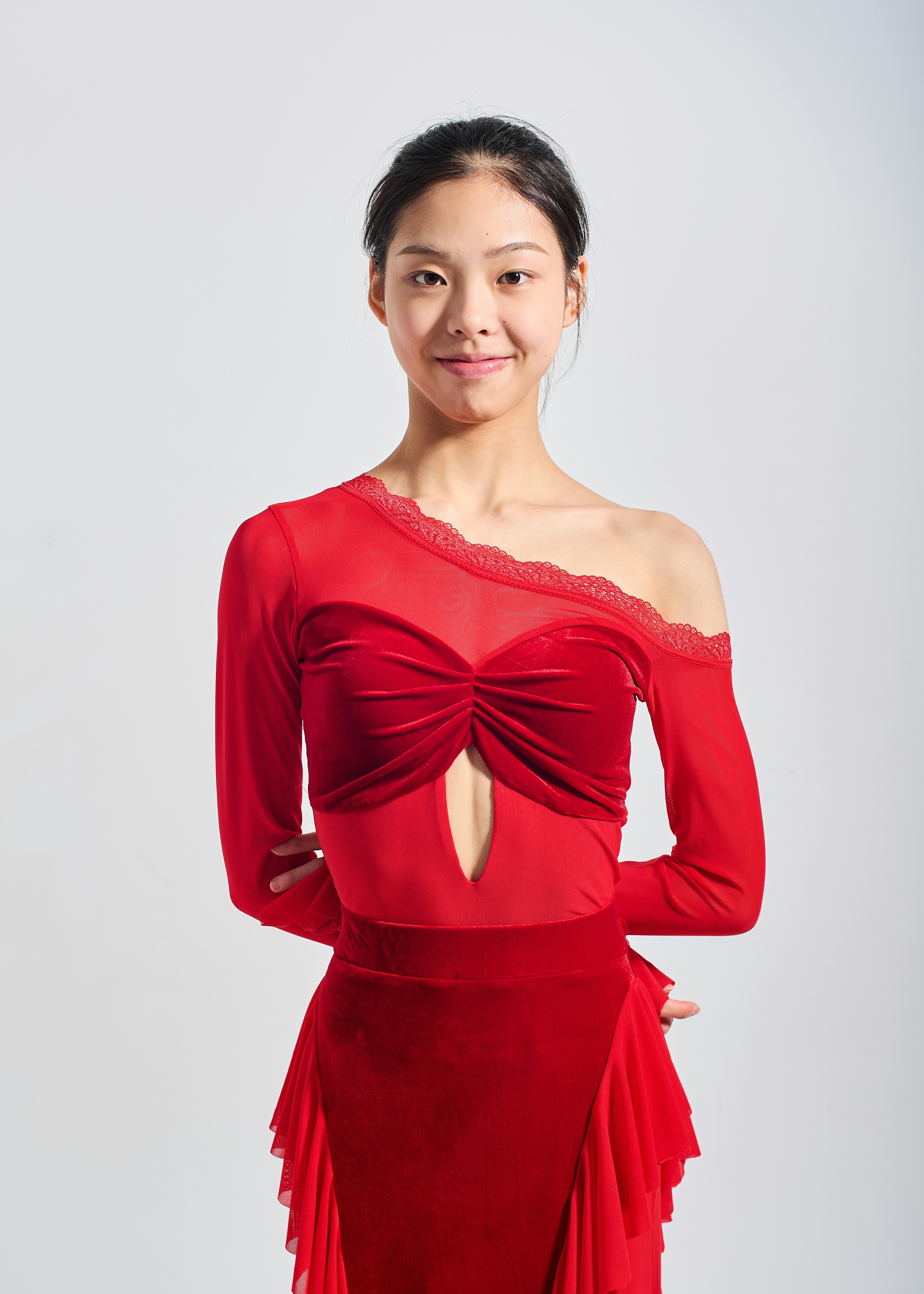 A poised dancer in a red outfit stands against a neutral backdrop, showcasing a single-shoulder design with ruching and a front cutout for dramatic silhouette. This studio shot emphasizes fabric texture and posture. 