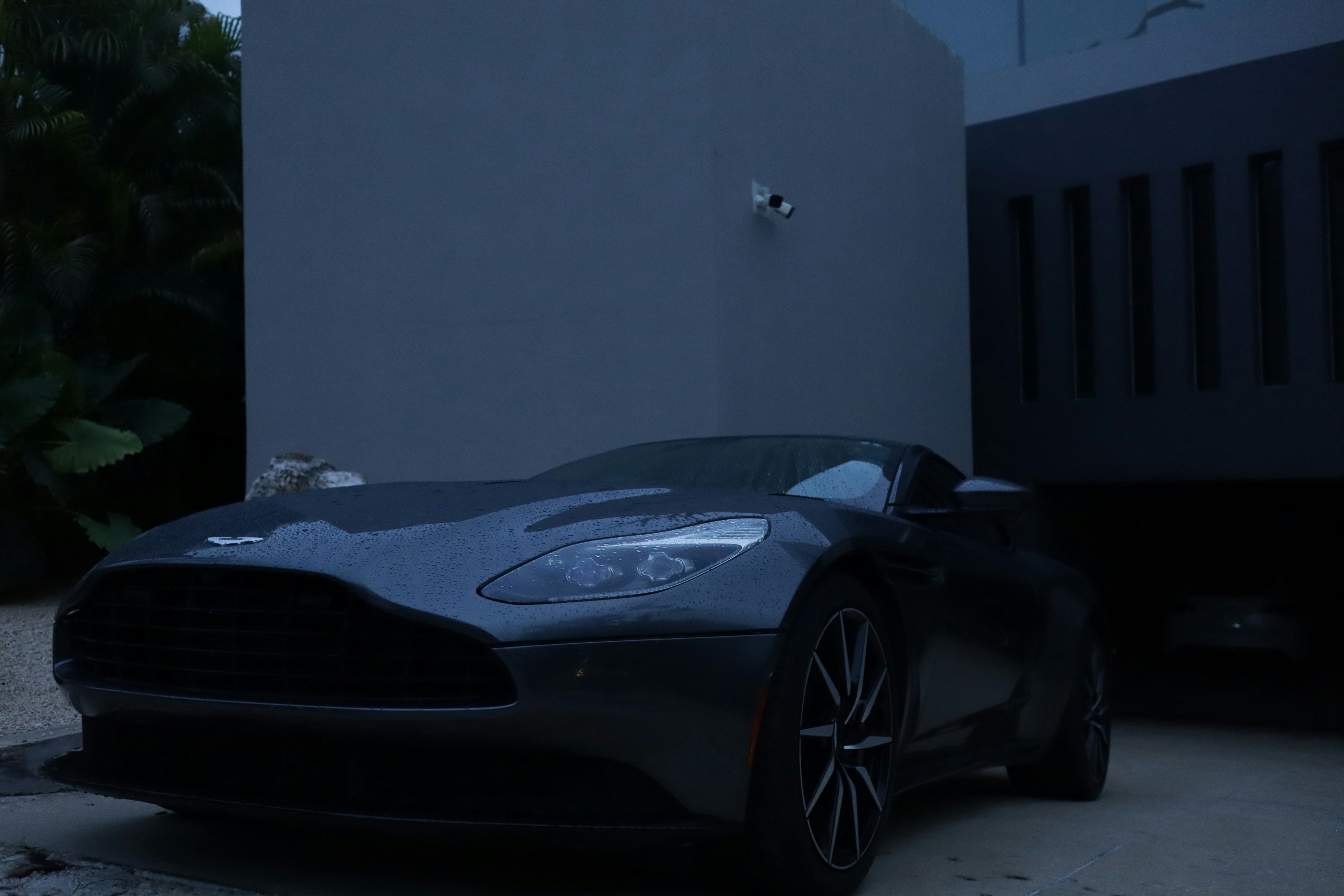 Dark-toned sports car parked in a shadowed driveway beside a modern building, with subtle reflections and dim outdoor lighting. This is a photograph-focused shot of a vehicle, not a painting or illustration.