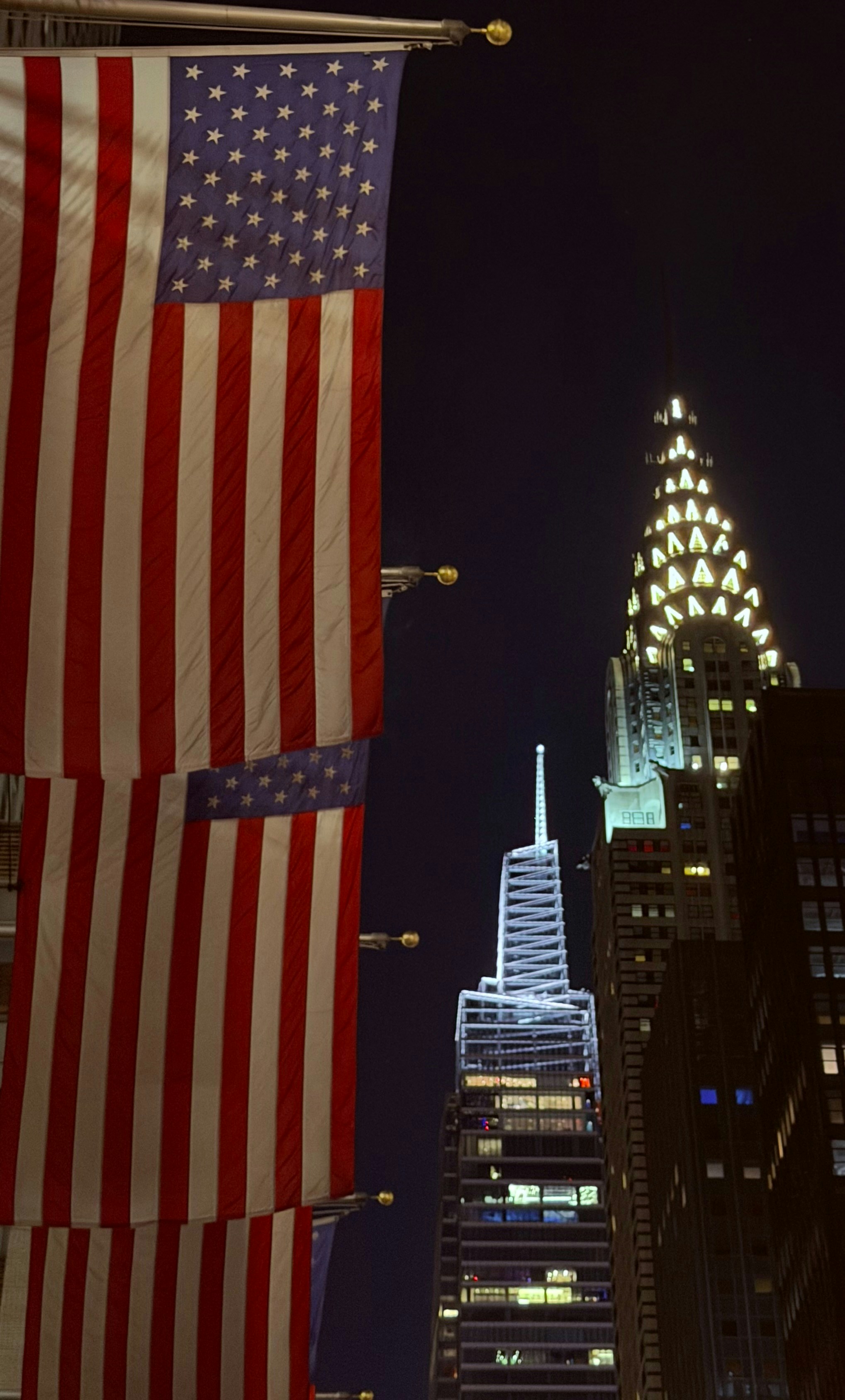 Close-night urban scene featuring a vertical American flag prominently on the left with illuminated skyscrapers in the background, showcasing a patriotic cityscape at night.